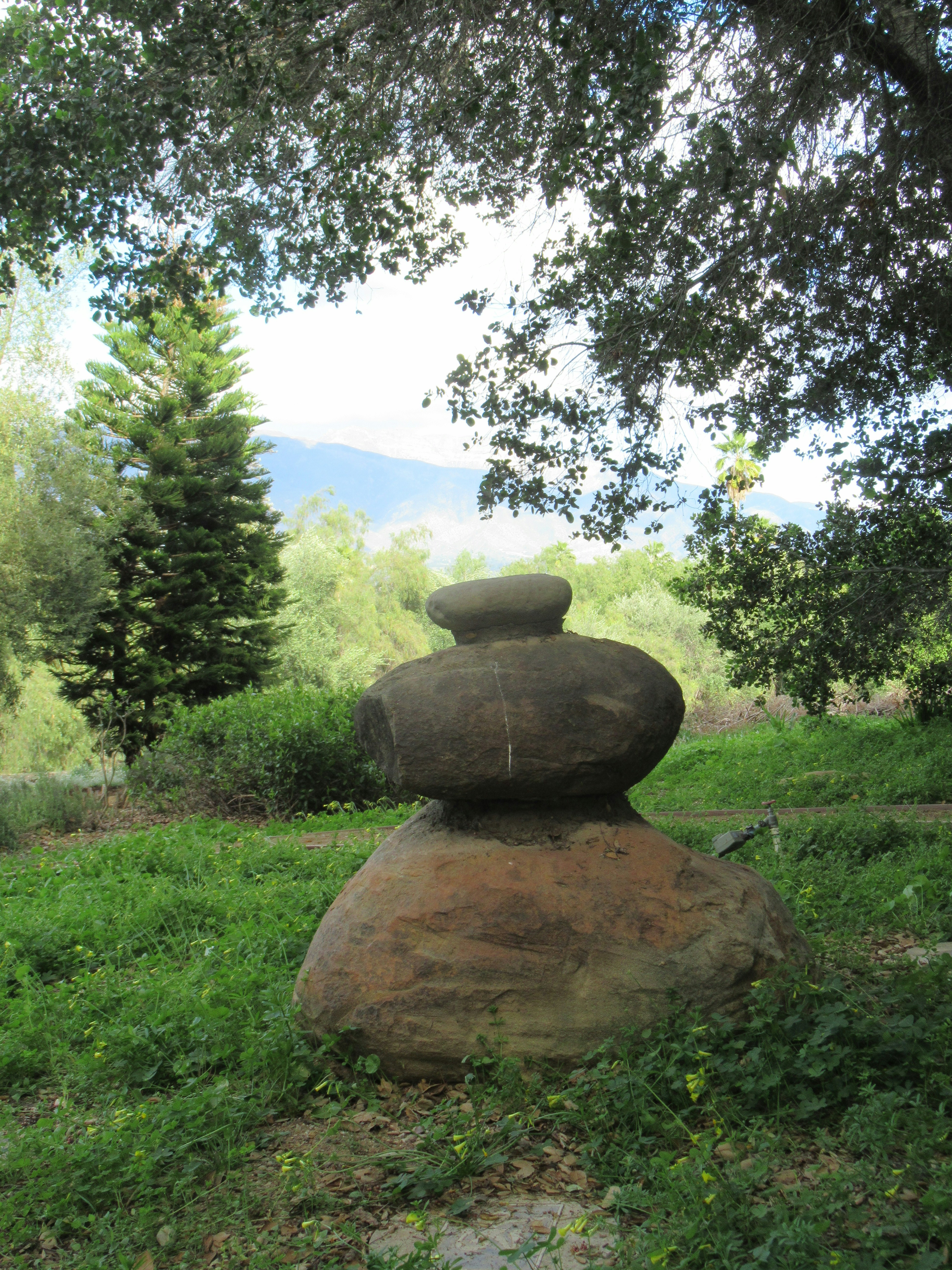 Stacked rock sculpture sits on a grassy patch beneath a leafy canopy, with distant hills in the background. The scene captures a tranquil outdoor moment in a park.