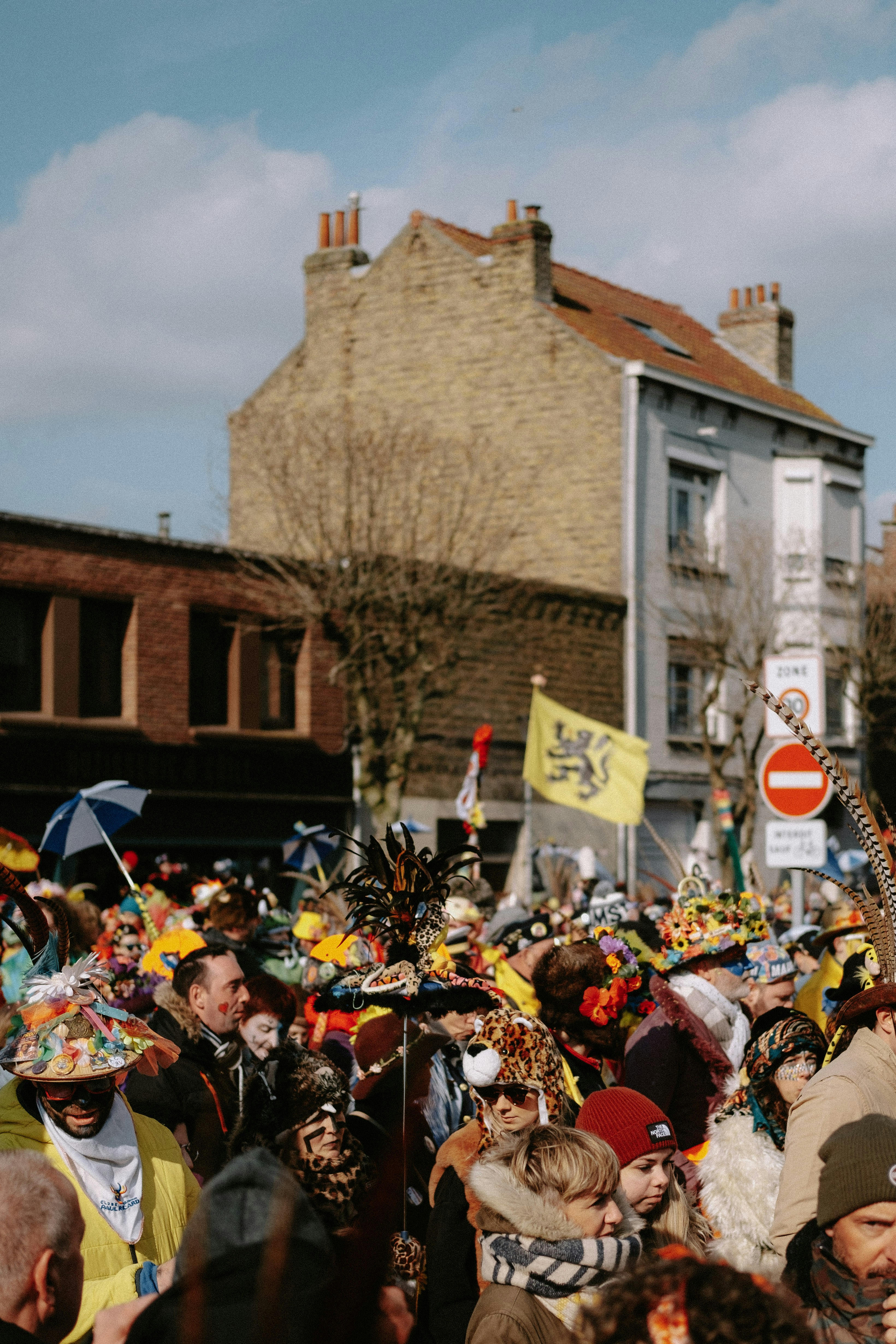 Crowd of revelers adorned in colorful costumes and hats during a festive parade, with buildings in the background. Flags flutter amidst the joyful atmosphere.