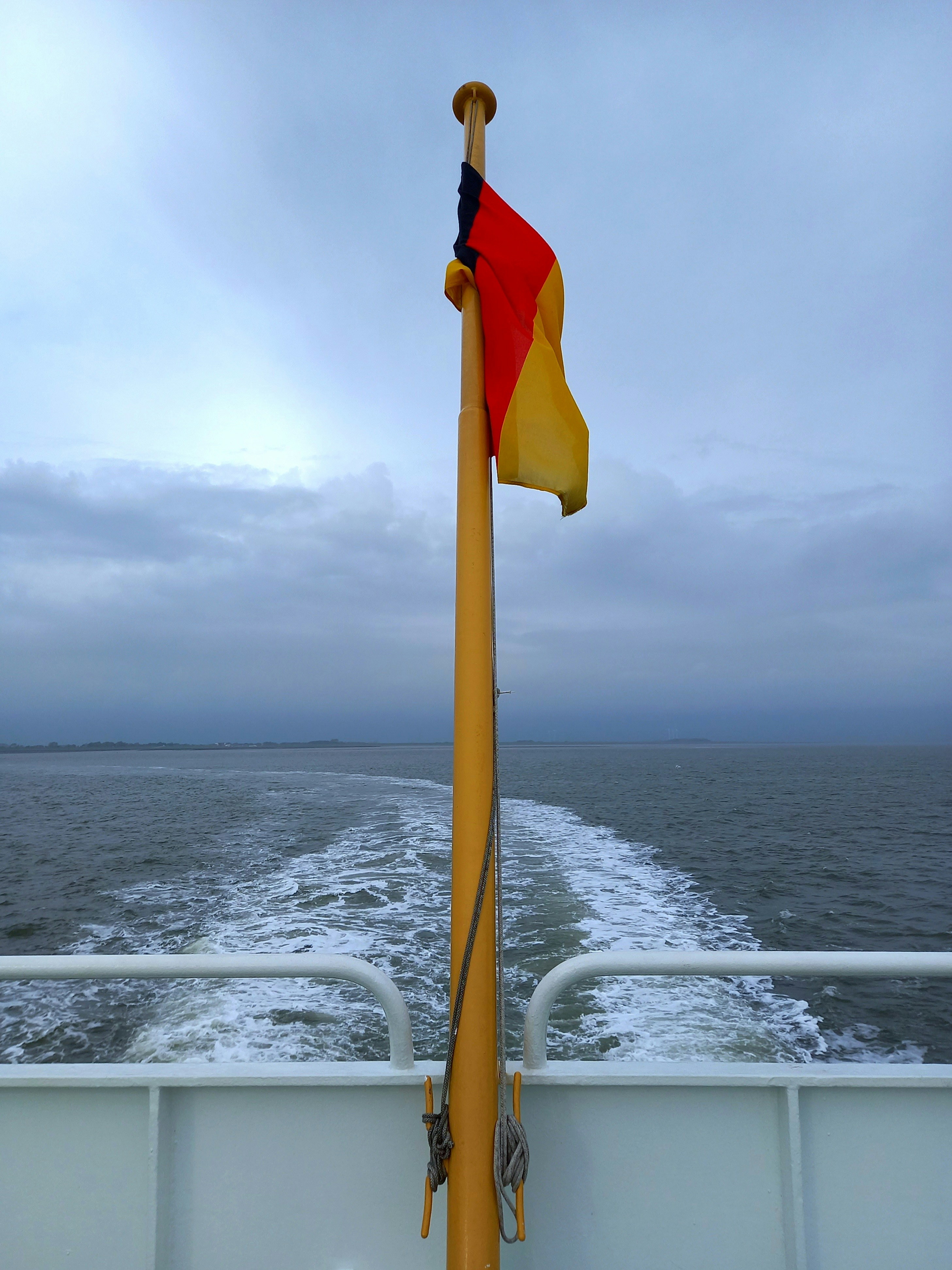 German flag fluttering atop a ship's mast with a wake trailing behind, set against a cloudy sky. 