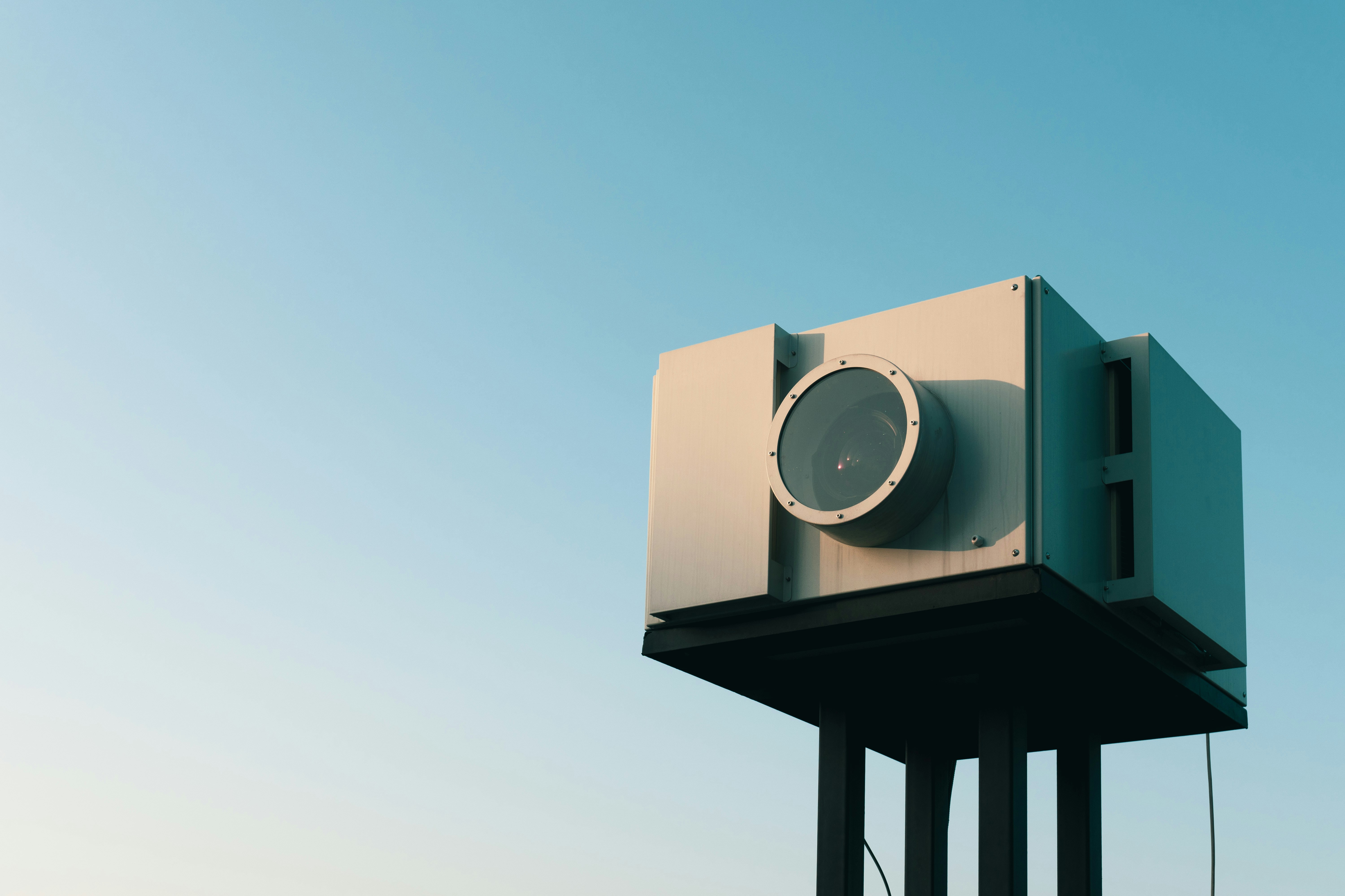 Roof-Top Sentinel: a boxed rooftop camera gazing into a pale blue sky