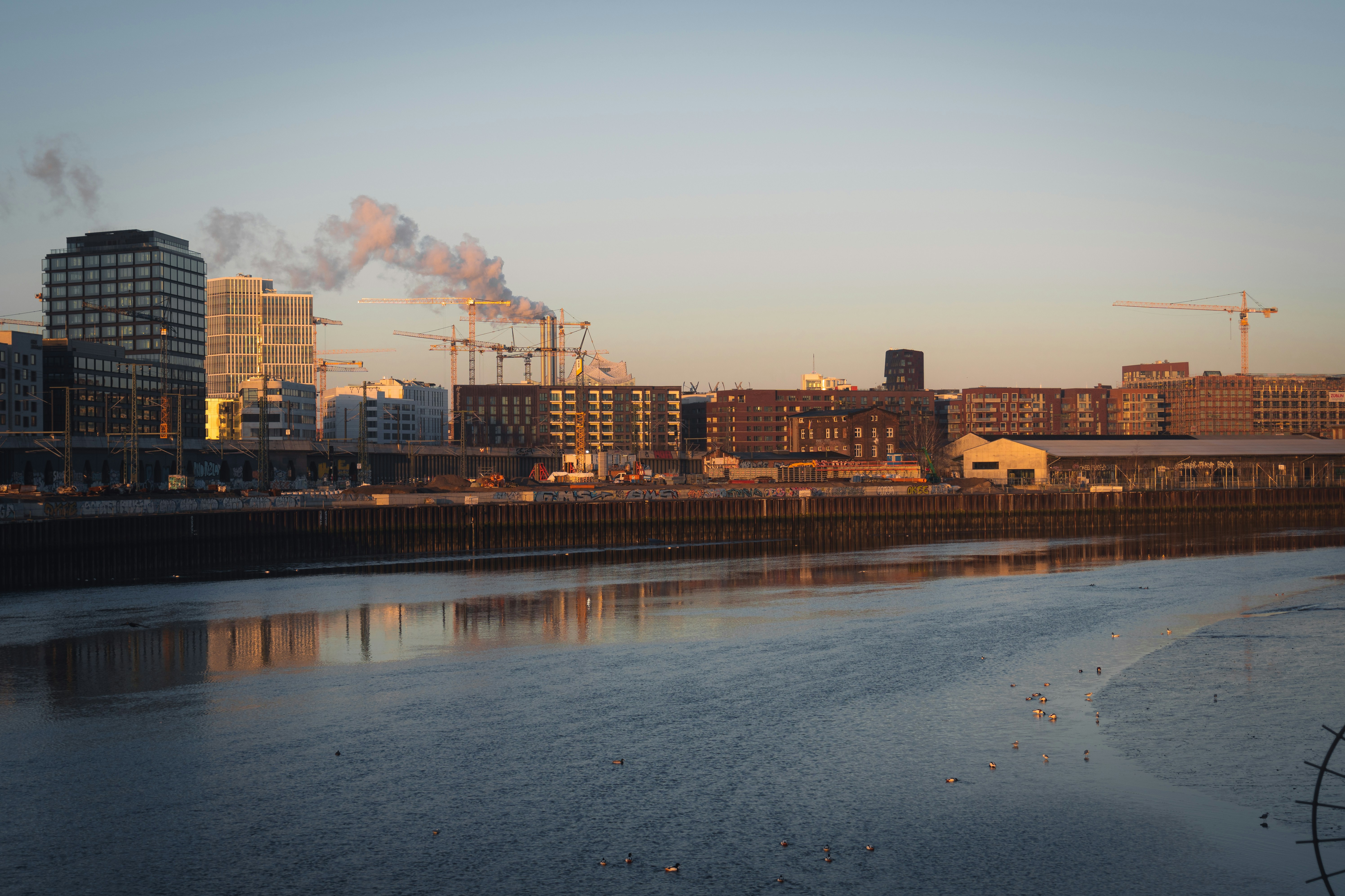 City skyline with smoke rising from chimneys, reflected on a tranquil river at sunset.