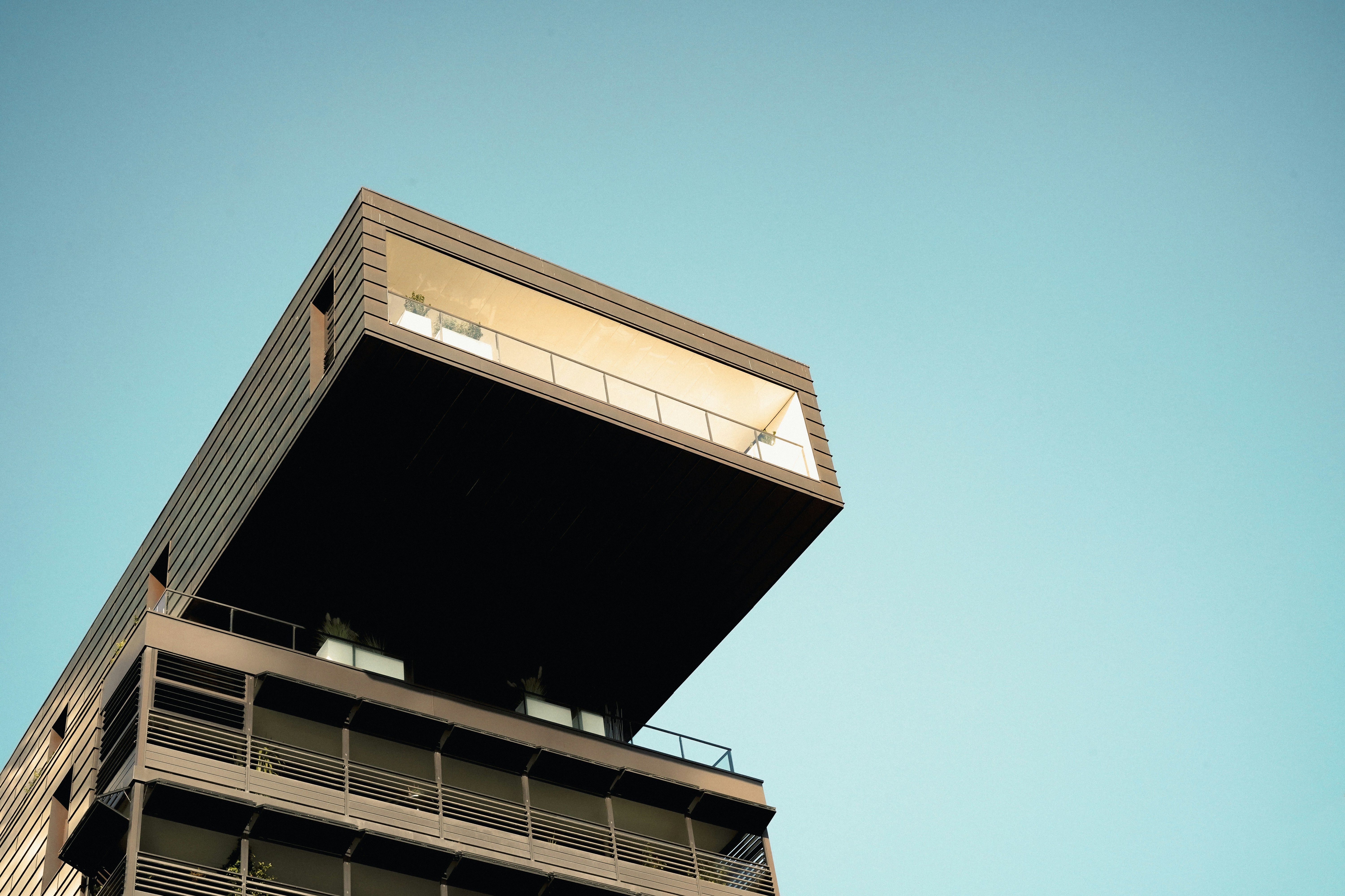 This striking image captures the angular design of a modern building jutting boldly against a pristine blue sky. The composition emphasizes the building's sleek lines and geometric forms, with warm sunlight bathing the upper floors and creating a stark contrast against the cool tones of the sky. The minimalist aesthetic and dynamic perspective make this photograph visually captivating and architecturally intriguing.