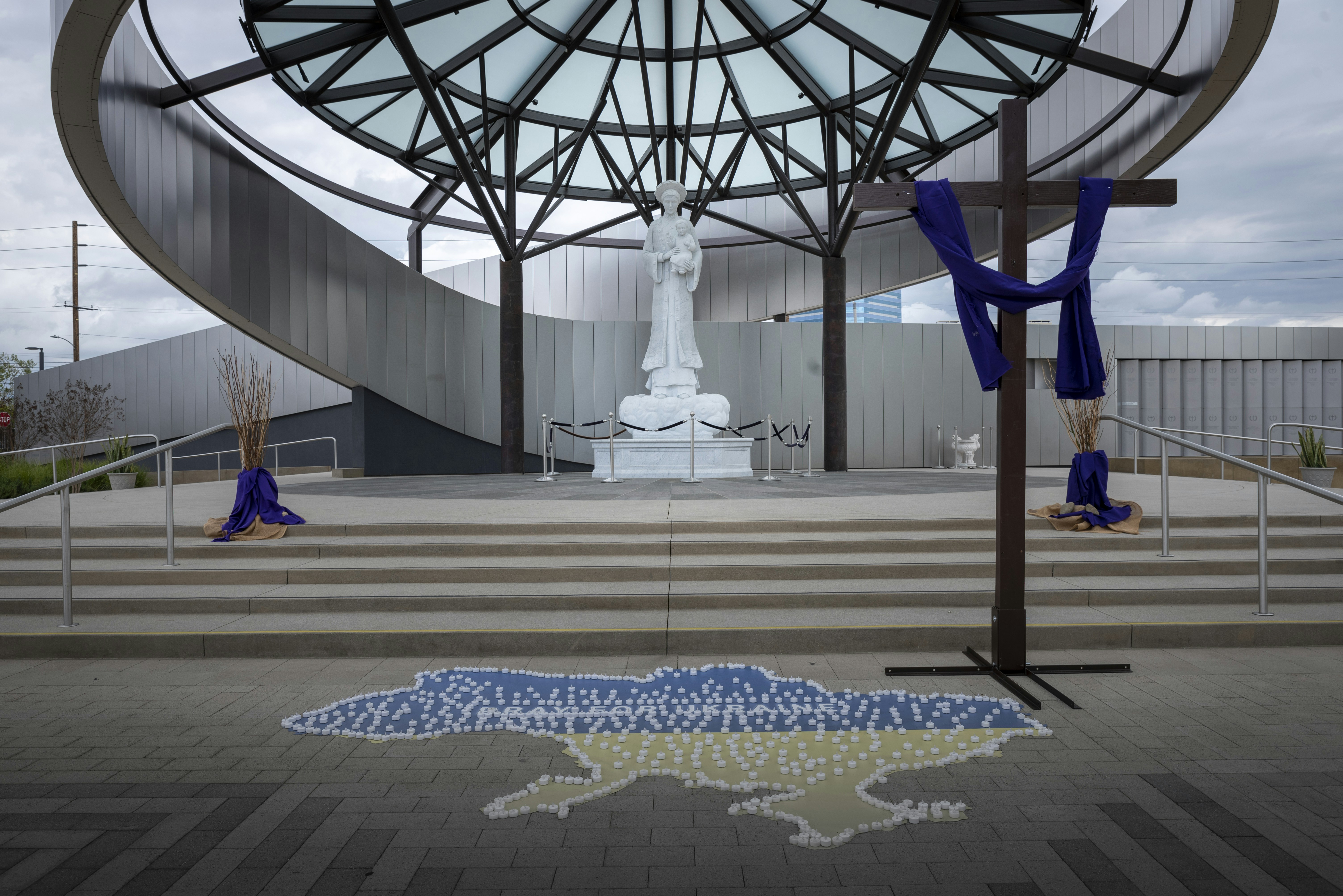 Two individuals perform yoga poses on steps beneath a circular modern canopy, with a large cross and statue in the background.