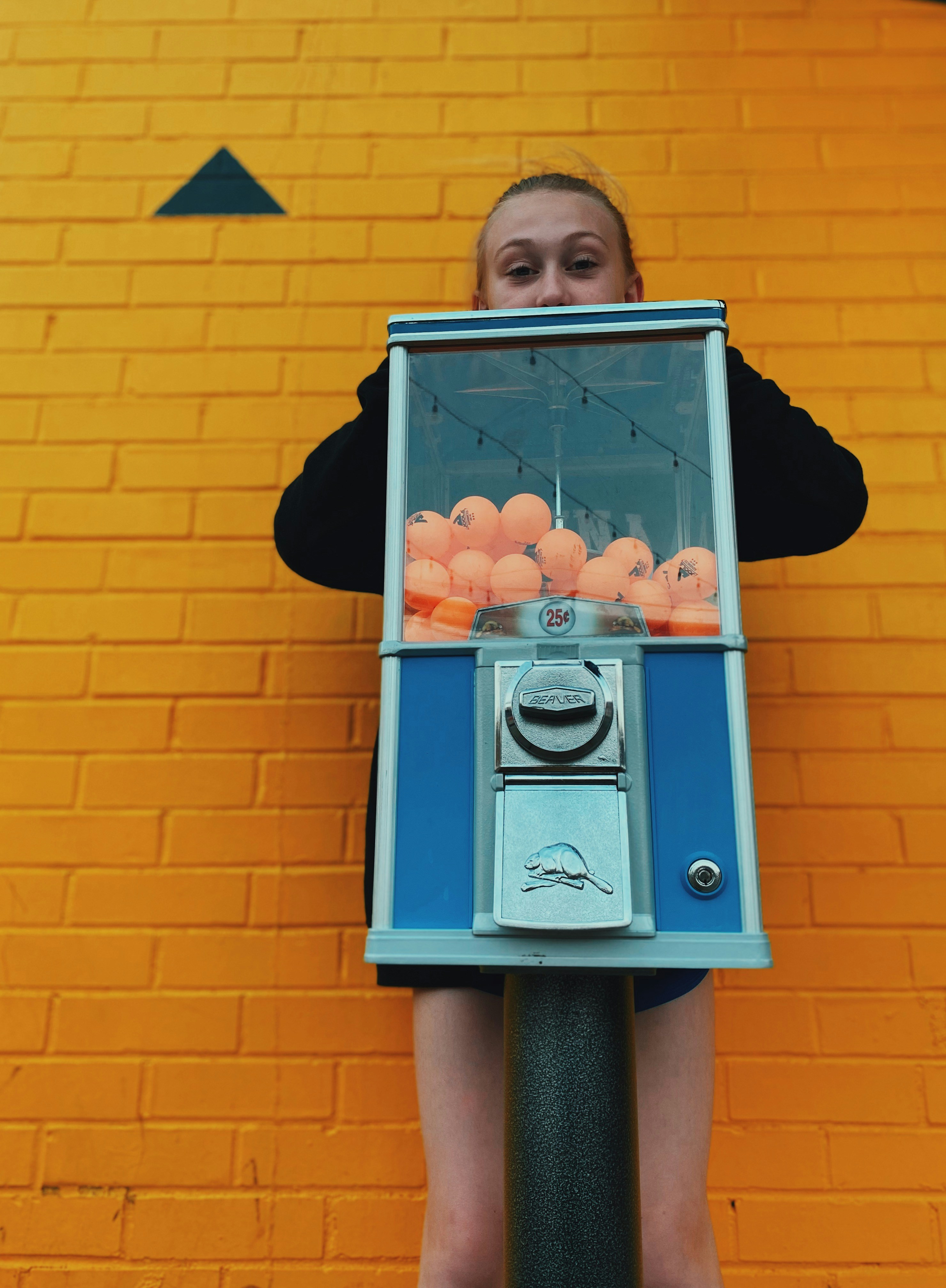 Blue Gumball Machine on a Pole Against a Neon Orange Brick Wall