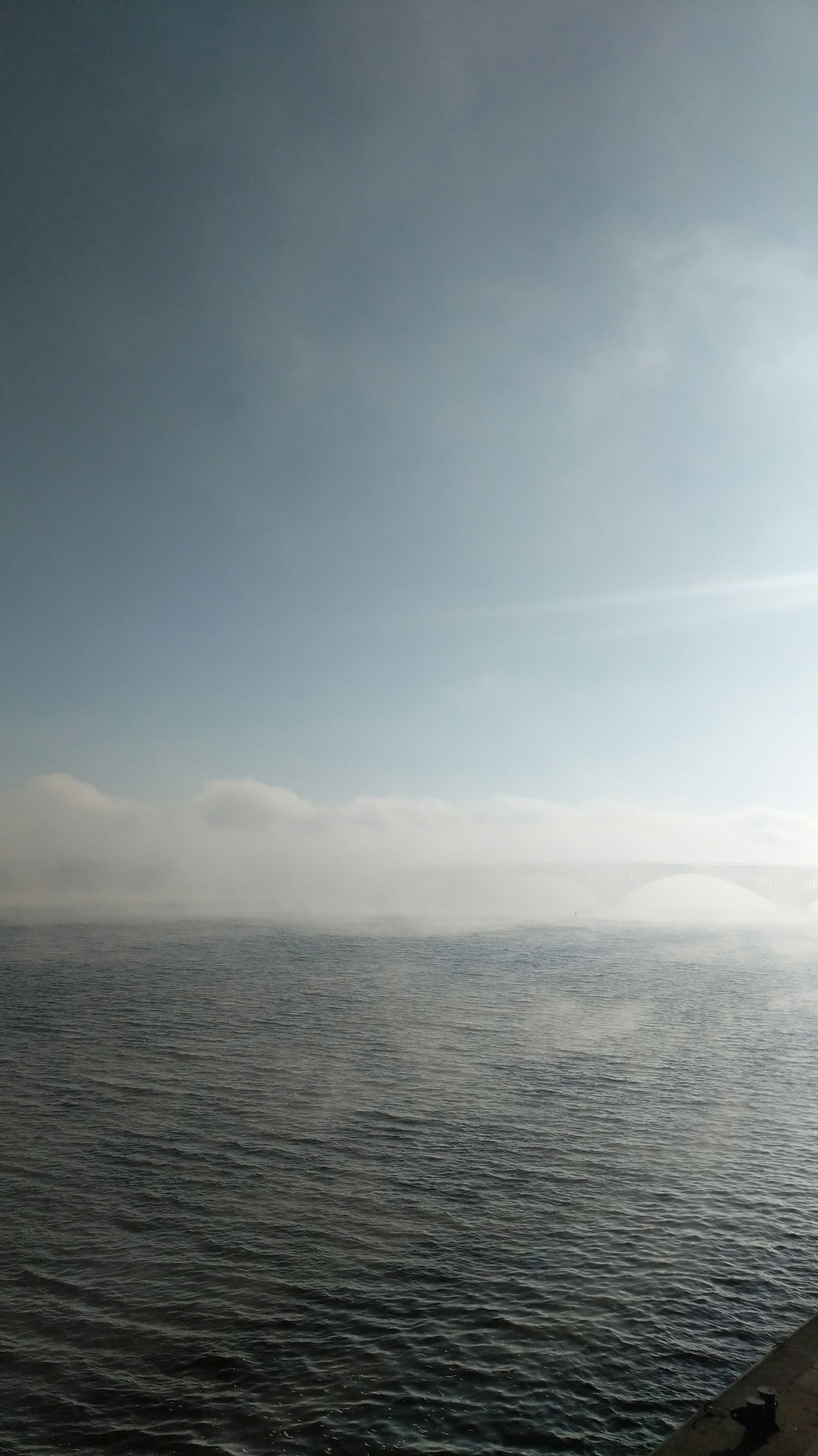 A tranquil seascape shrouded in mist, with soft waves reflecting the pale sky. The horizon is blurred, creating a dreamlike atmosphere.