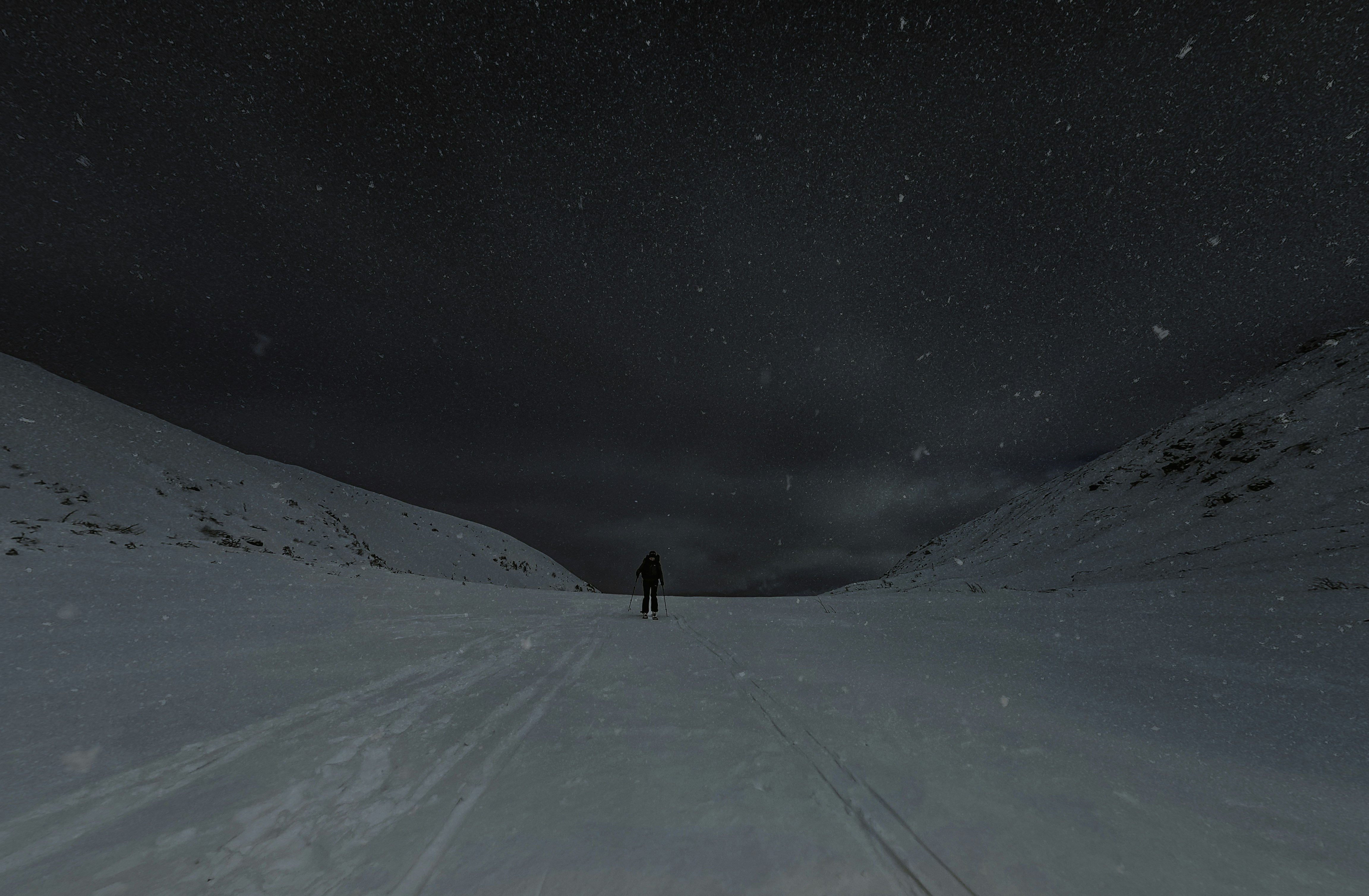 A lone figure stands on a snow-covered path beneath a star-filled night sky, framed by dark, rolling hills.