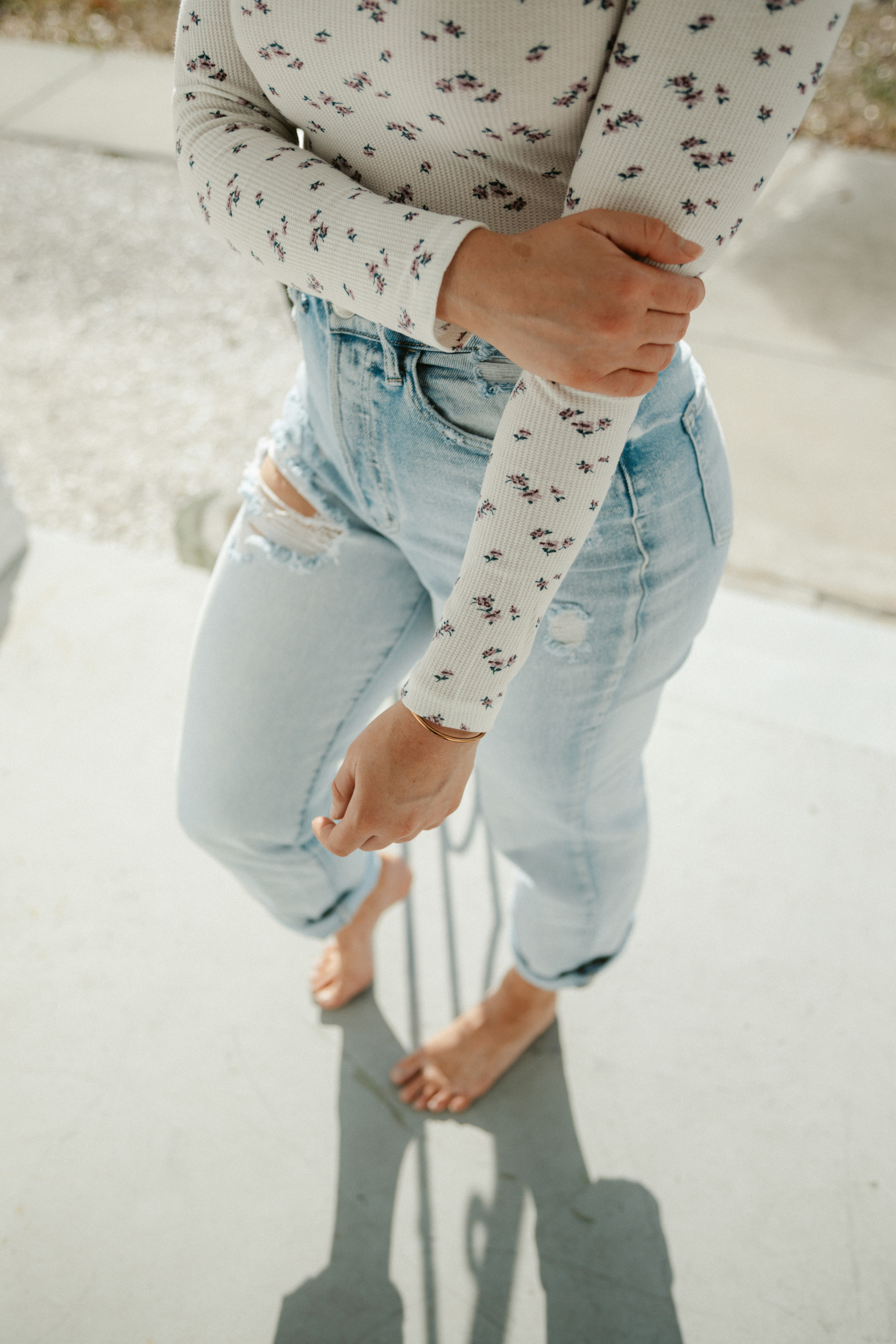 A young woman in a floral long-sleeve top and distressed jeans stands barefoot on a sunlit porch, showcasing a relaxed yet stylish vibe.