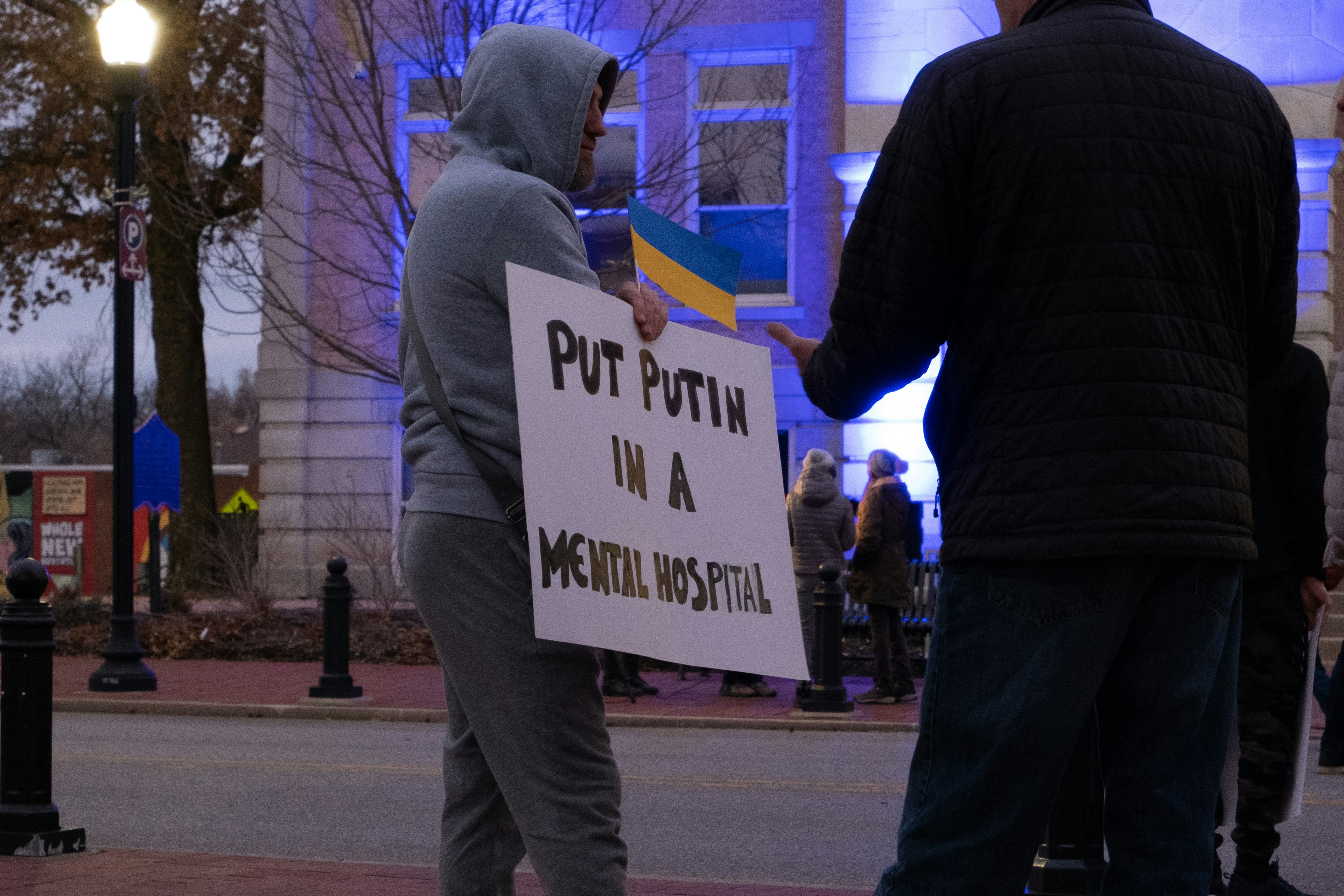 Protester holding a sign demanding action against Putin, with a Ukrainian flag in the background. The scene captures the essence of public outcry and activism.