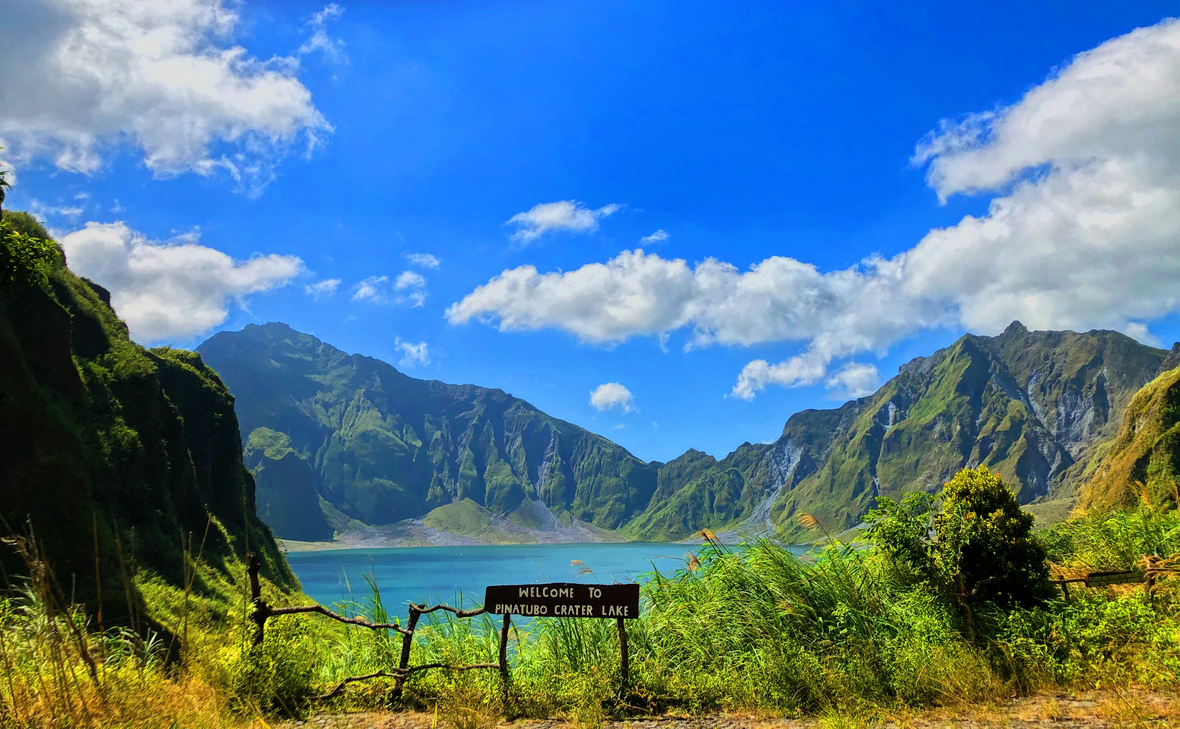 Lush greenery frames a serene crater lake surrounded by rugged mountain peaks under a vibrant blue sky.