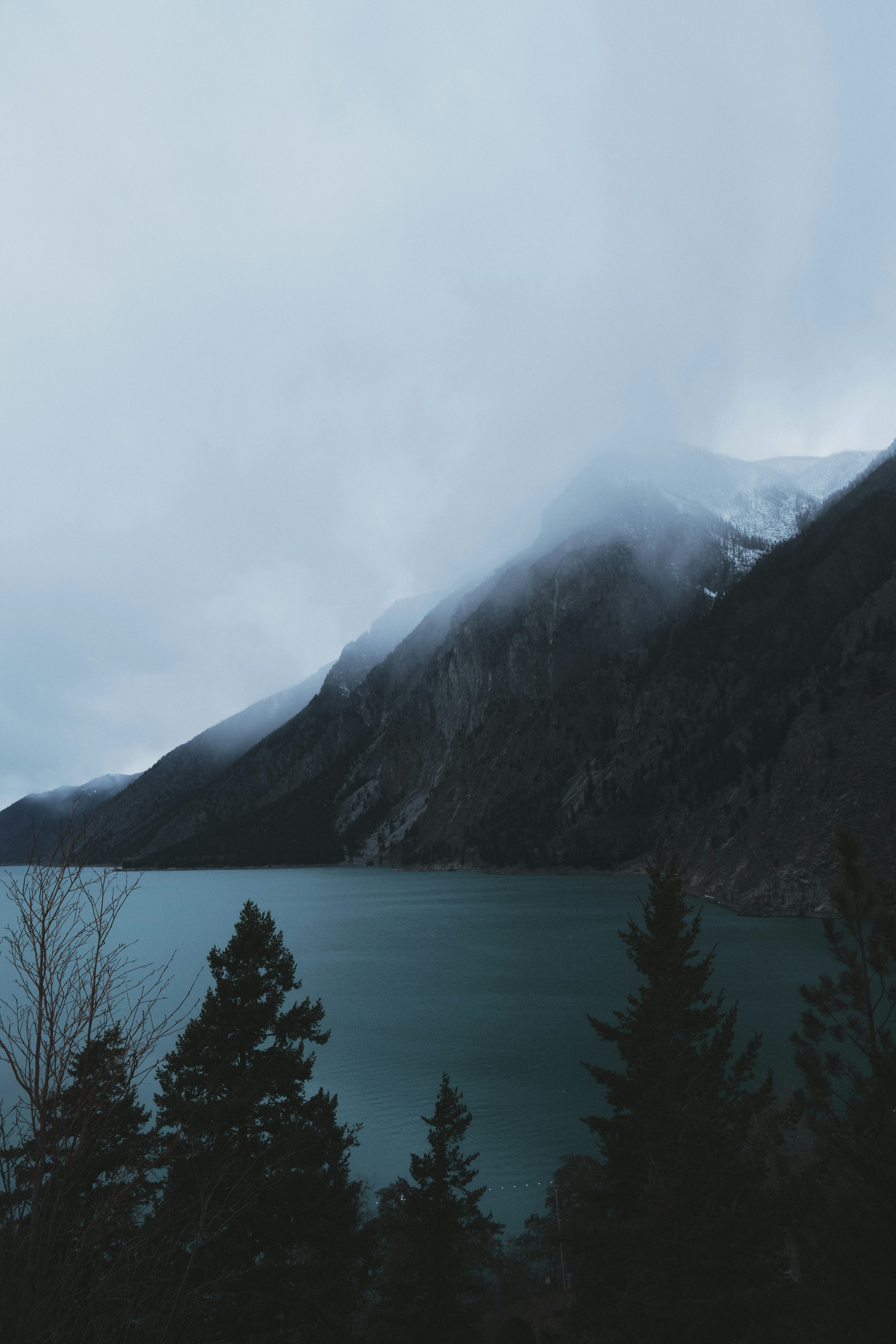 Misty mountains loom over a tranquil lake, framed by dark evergreens, creating a serene, atmospheric landscape.