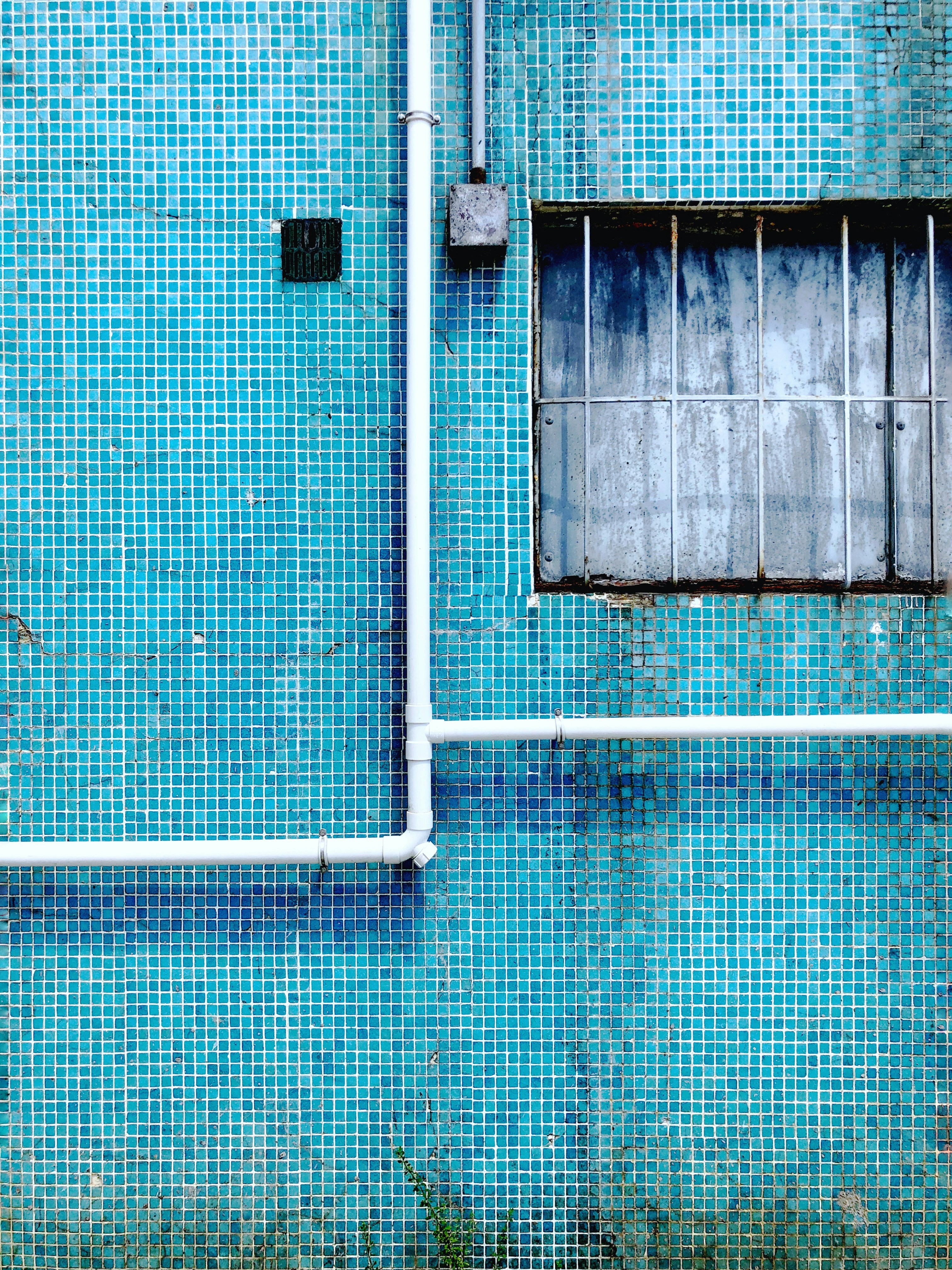 Textured blue tiled wall featuring white piping and a barred window, illustrating urban decay and color contrast.