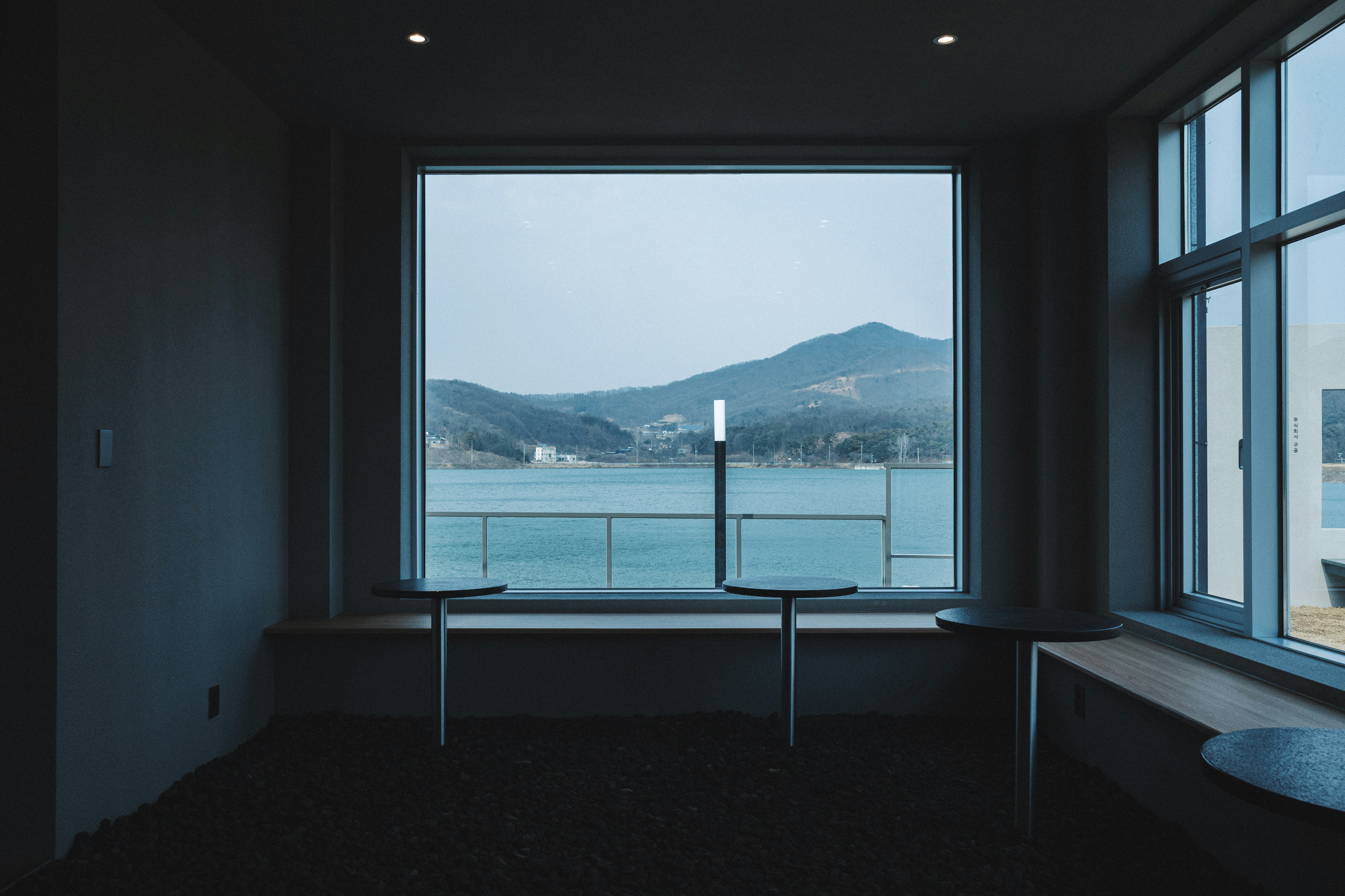 Serene interior space featuring a large window overlooking a calm harbor and distant mountains, with minimalist furniture. 