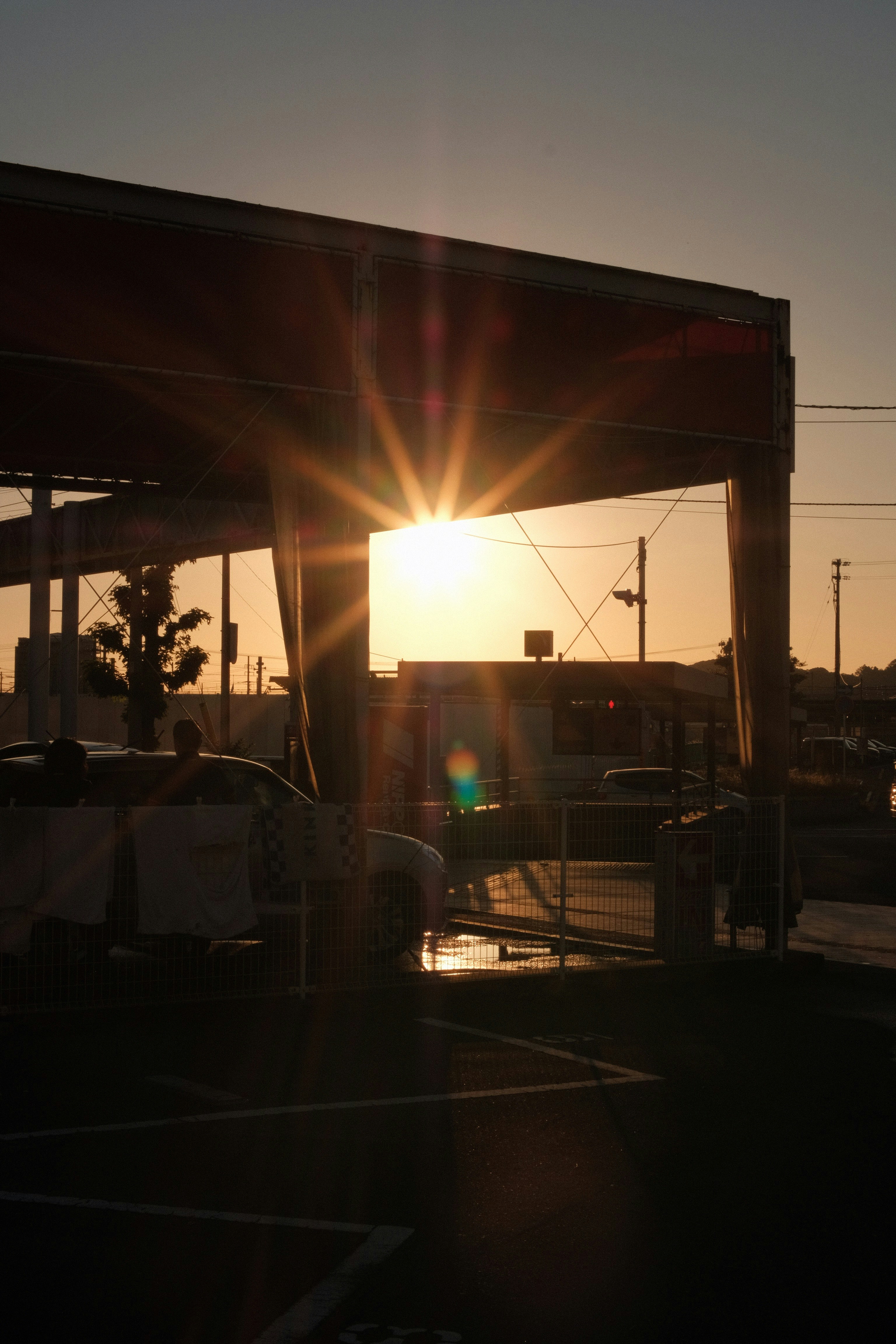 Golden sun setting behind a canopy, casting rays and creating silhouettes of nearby structures. The warm glow highlights the transition from day to night.