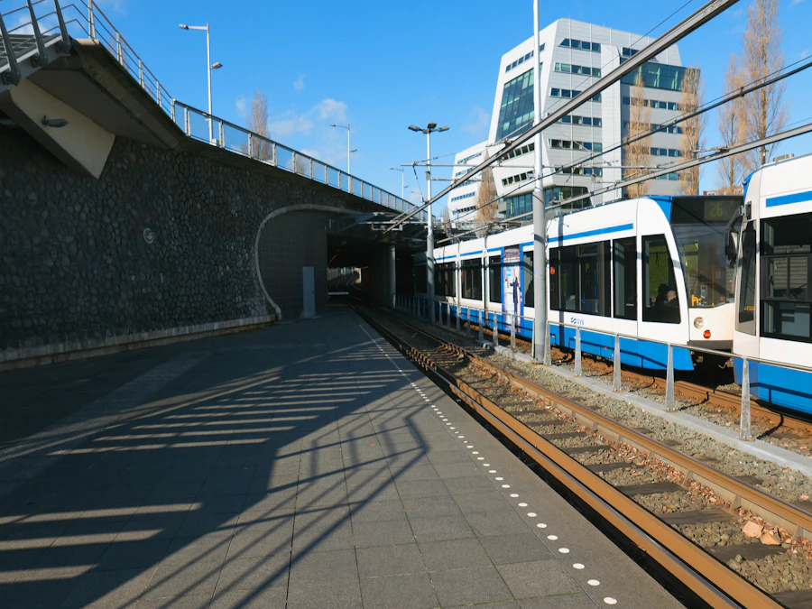 White-and-blue light rail trains curve past a platform beside a shadowed underpass, with office buildings rising behind the tracks under a clear blue sky.