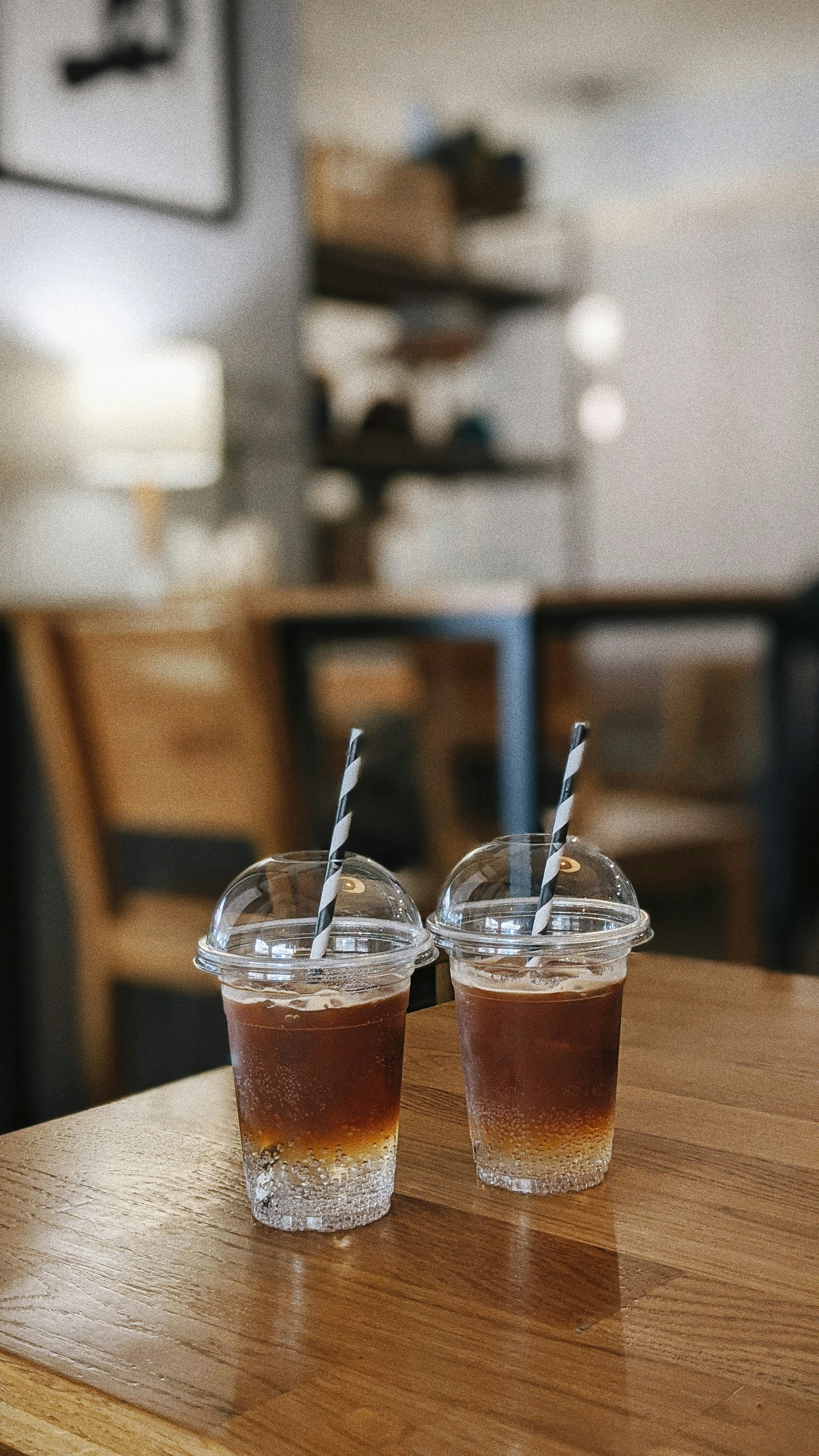Two iced beverages with striped straws resting on a wooden table in a cozy café setting.