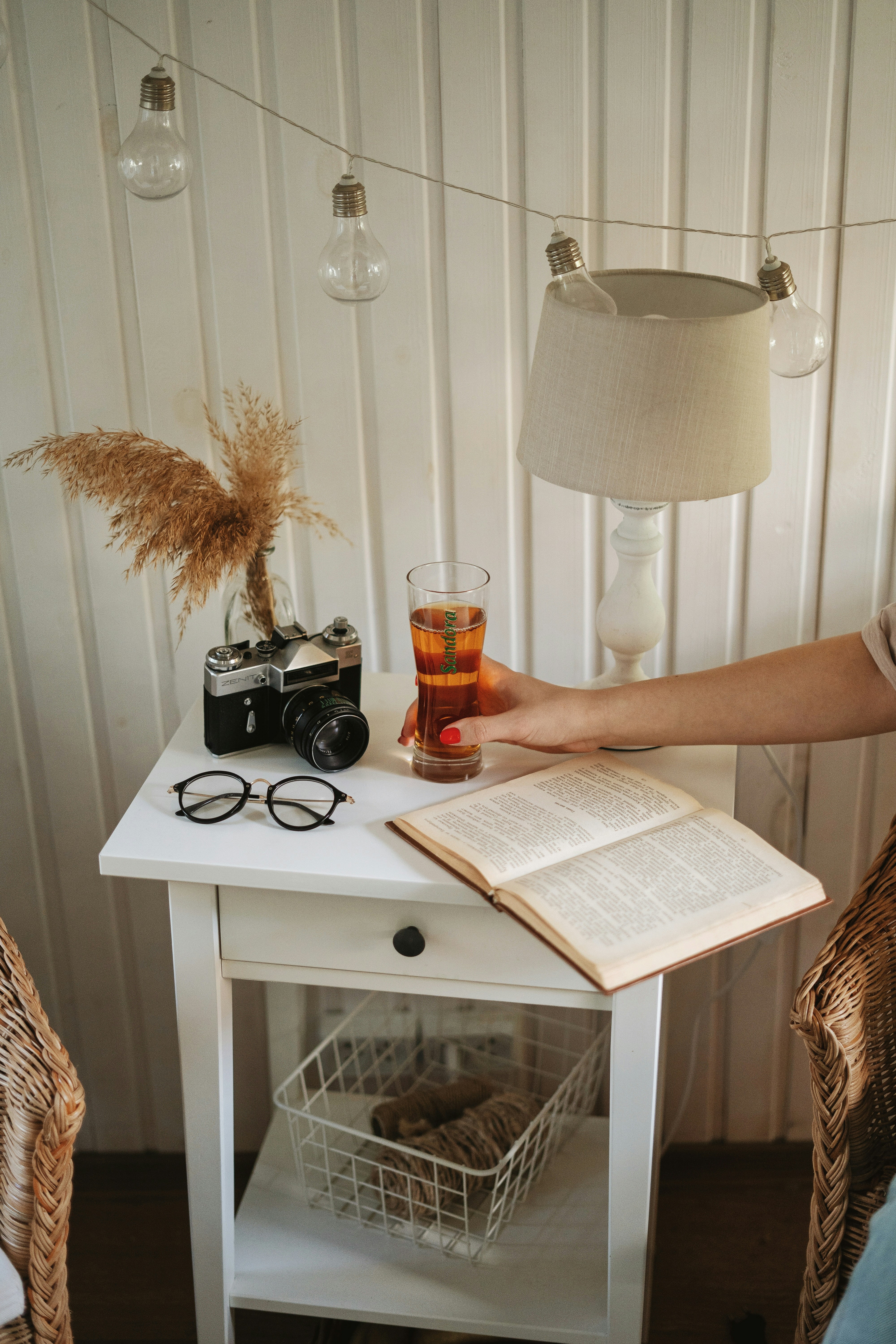 A vintage camera rests next to a glass of iced tea on a white side table, accompanied by a book and decorative elements. Soft lighting enhances the warm ambiance.