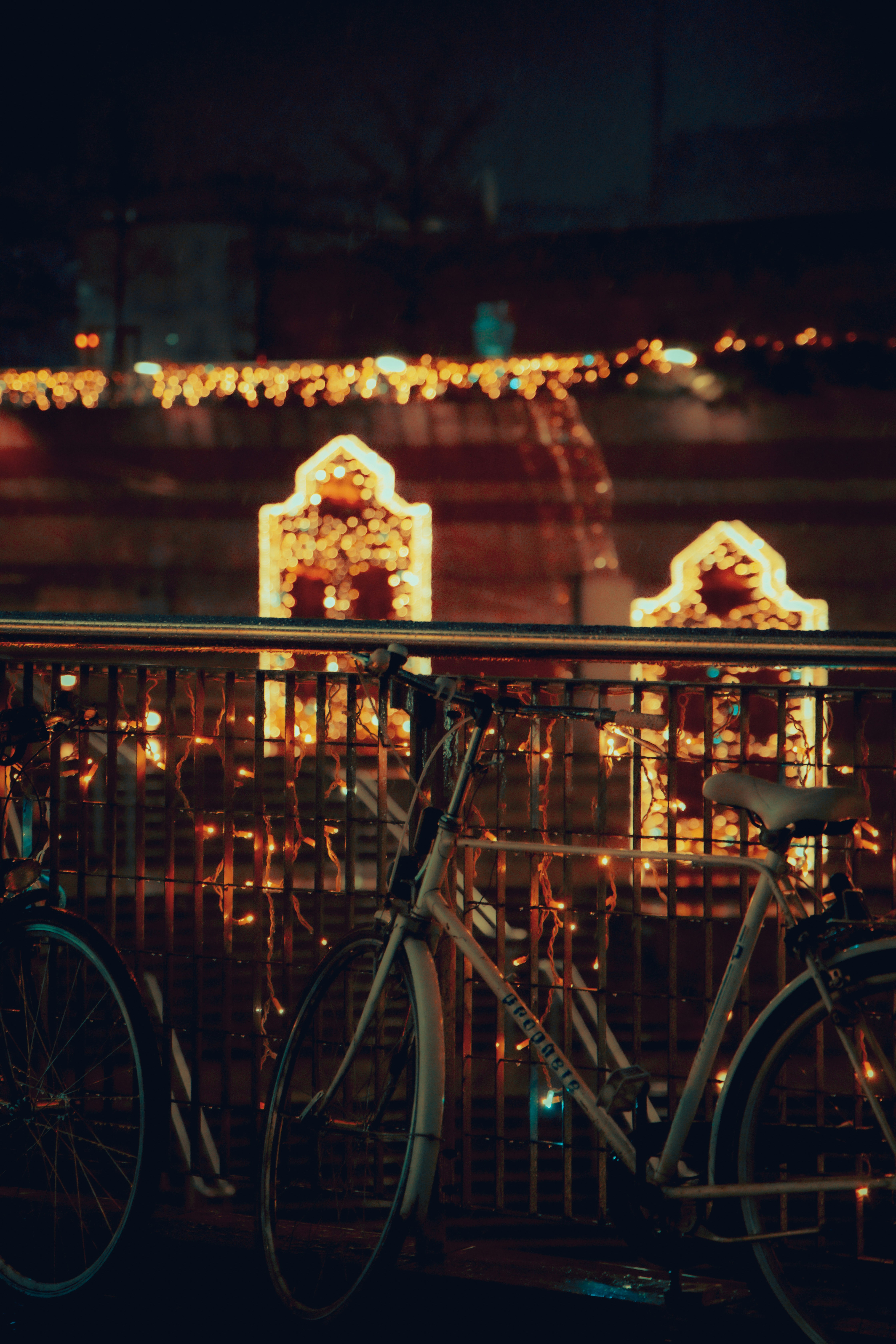 Bicycles rest against a railing, framed by twinkling lights and architectural details in the background. The scene captures a serene urban moment at night.