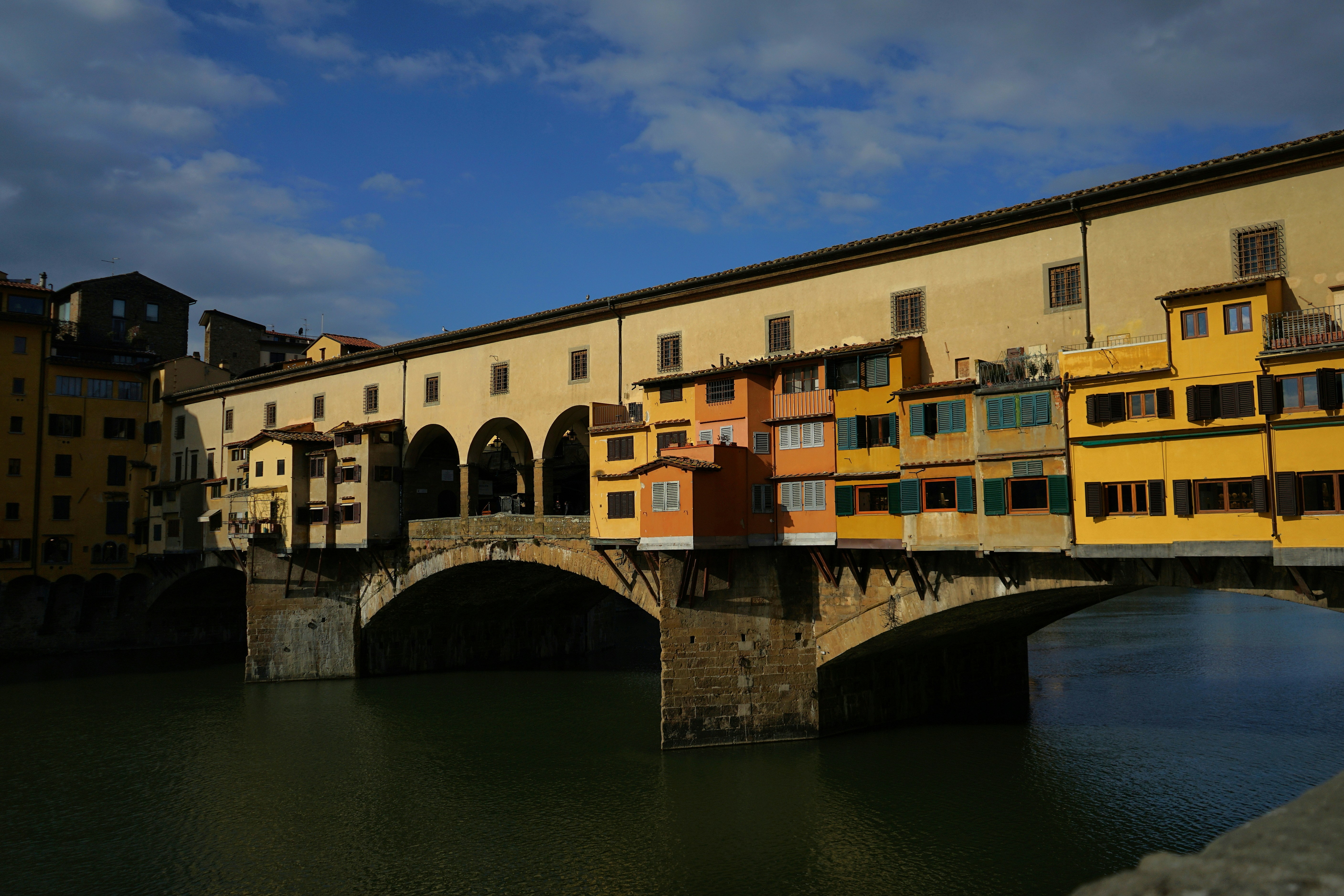 Ponte Vecchio adorned with colorful shops, arching over the Arno River under a partly cloudy sky.