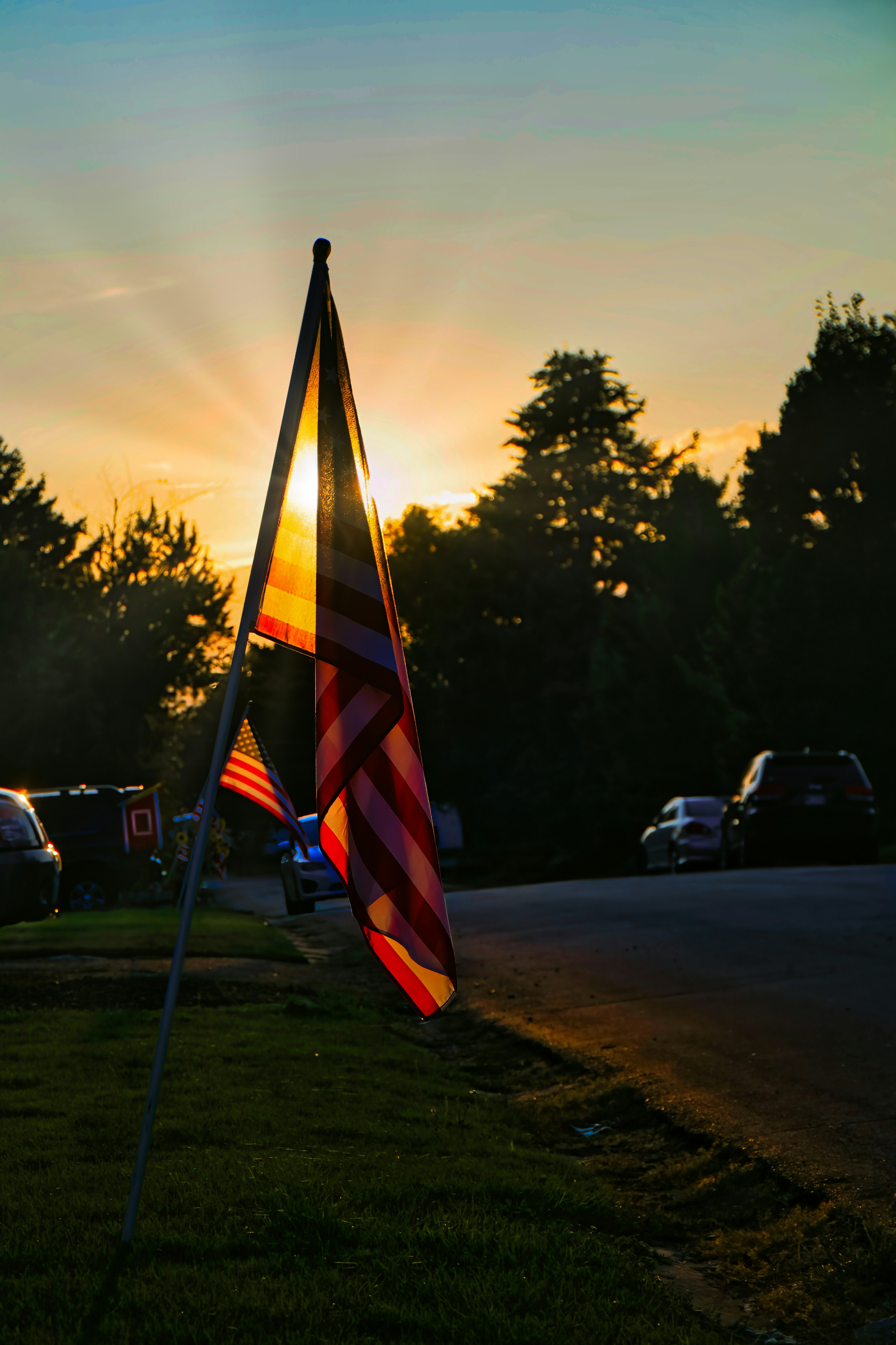 American flag illuminated by the setting sun, casting a warm glow against a backdrop of trees and distant vehicles.