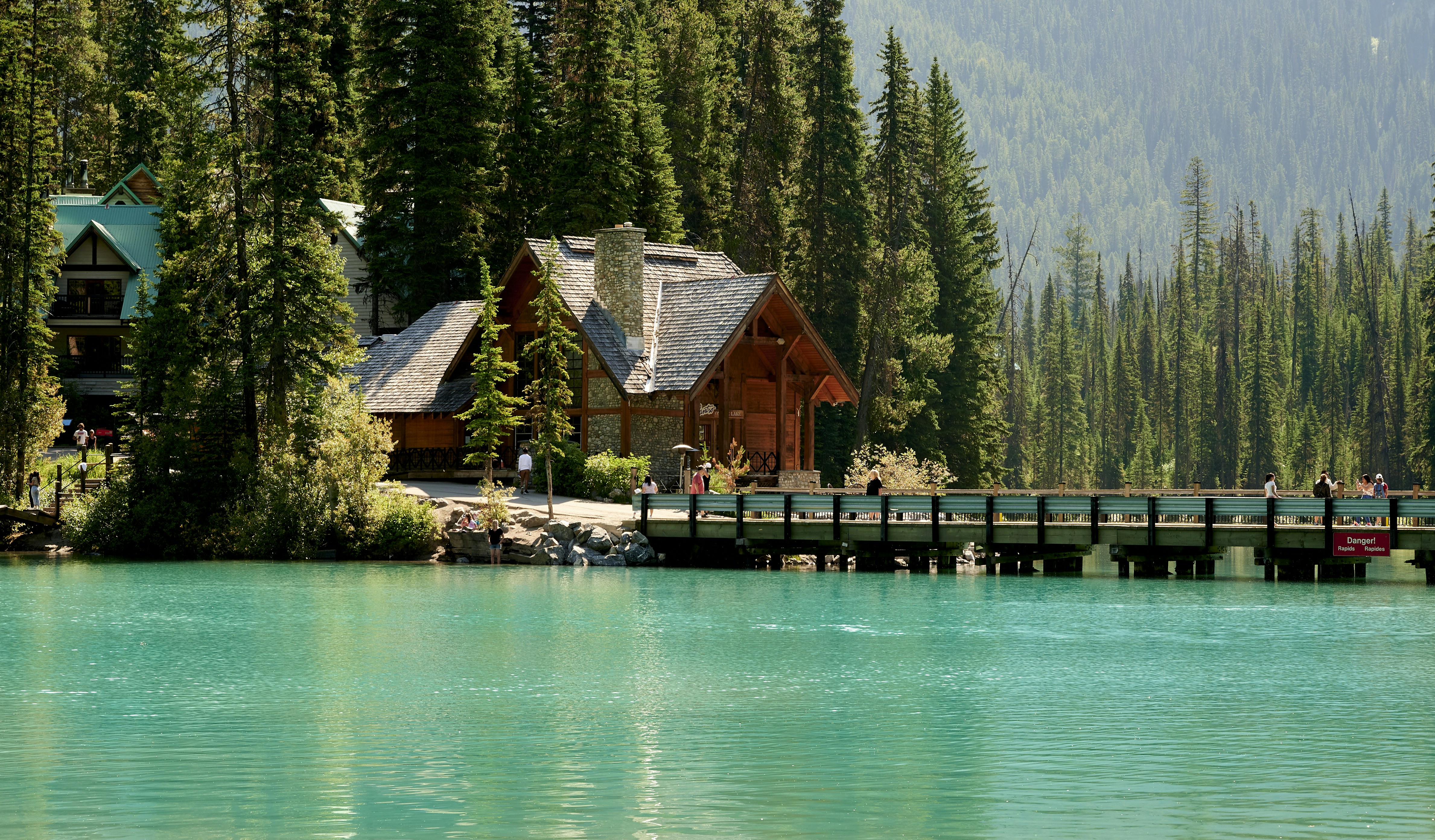 Charming wooden cabin nestled among lush pine trees beside a tranquil turquoise lake, with a wooden dock extending into the water.