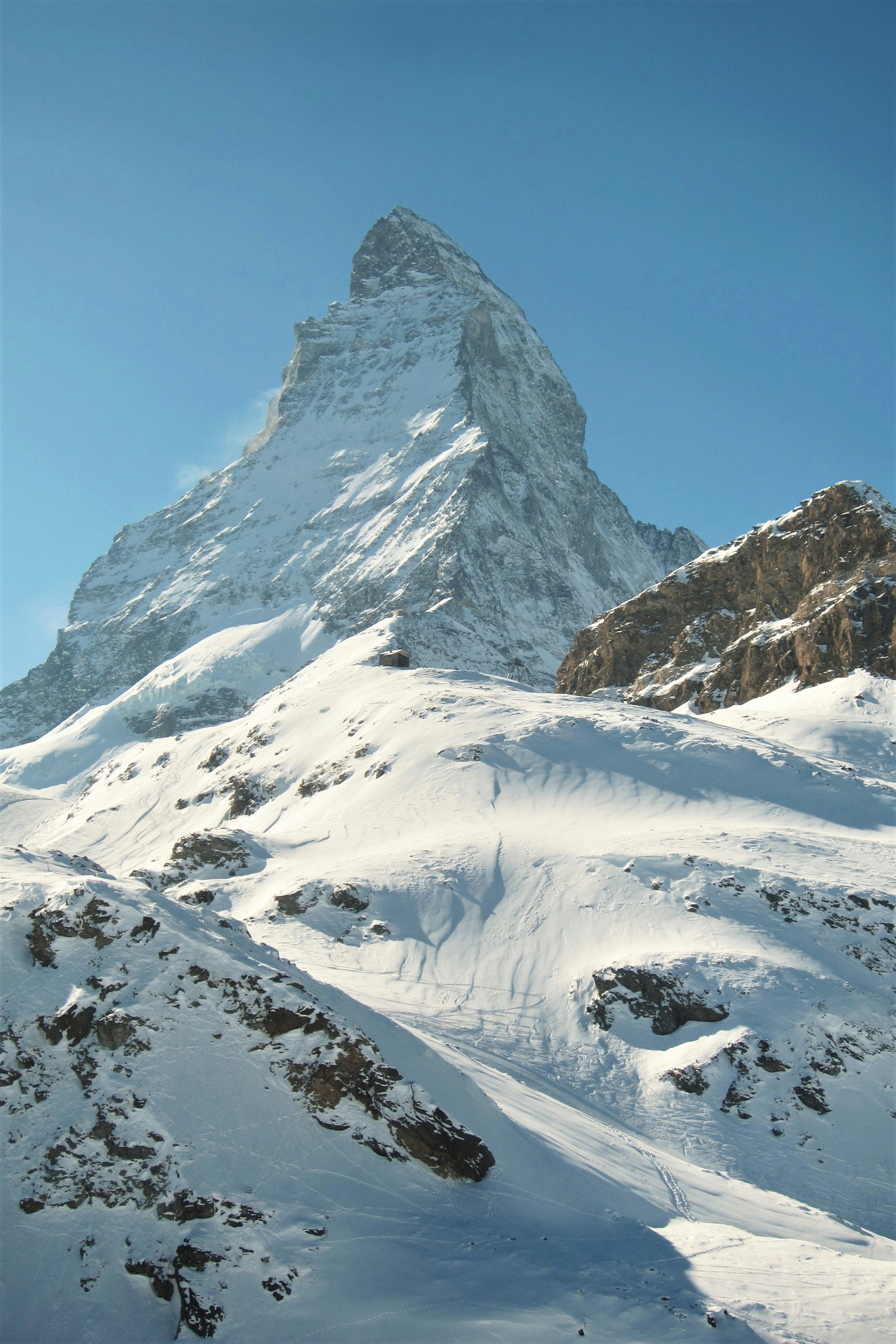 Photograph of a snow-covered alpine peak under a clear blue sky, with wind-sculpted ridges and smooth foreground snow.