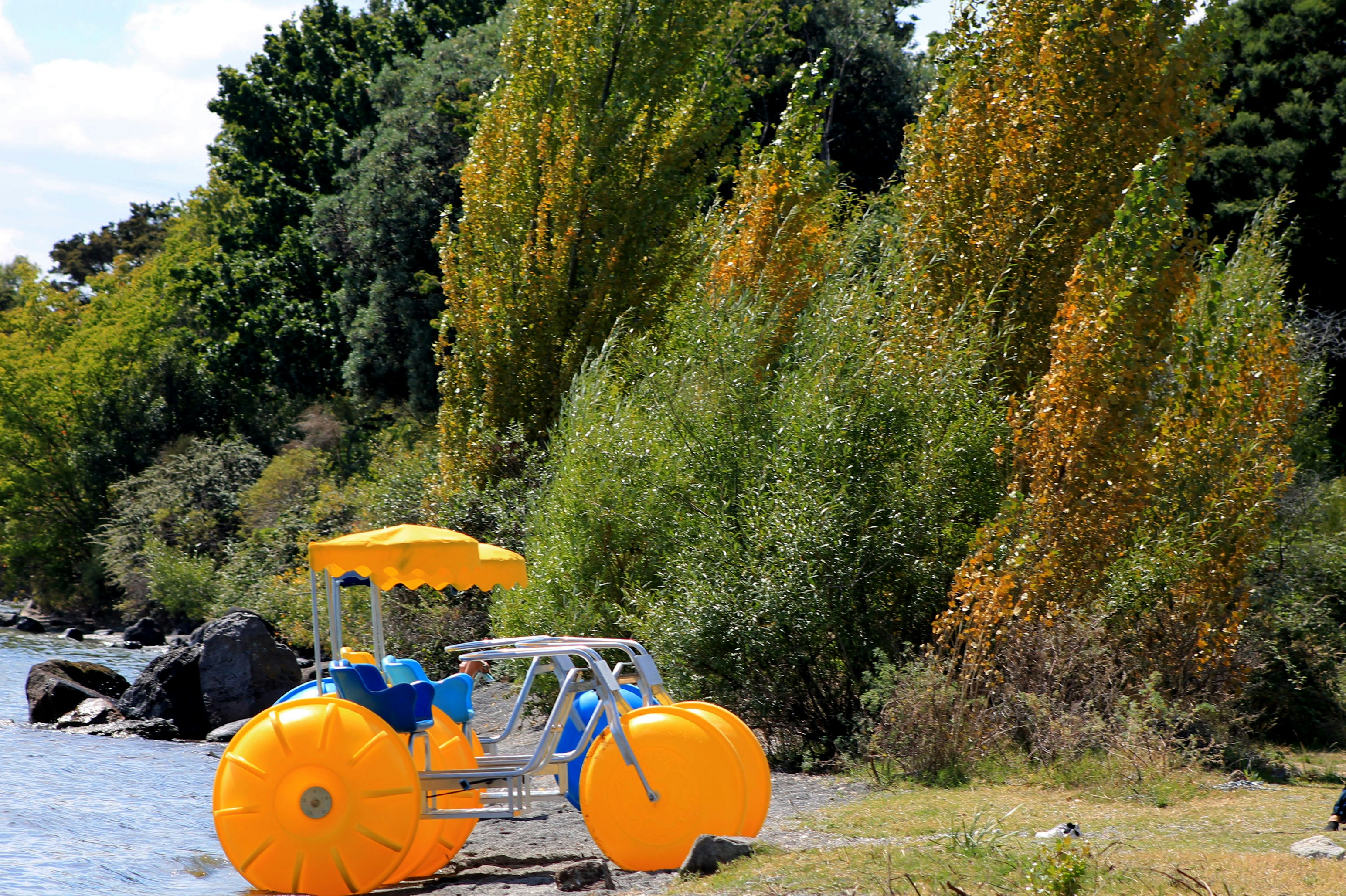Bright yellow pedal boat resting by the riverbank, surrounded by lush greenery and colorful trees. A perfect spot for leisurely summer outings.