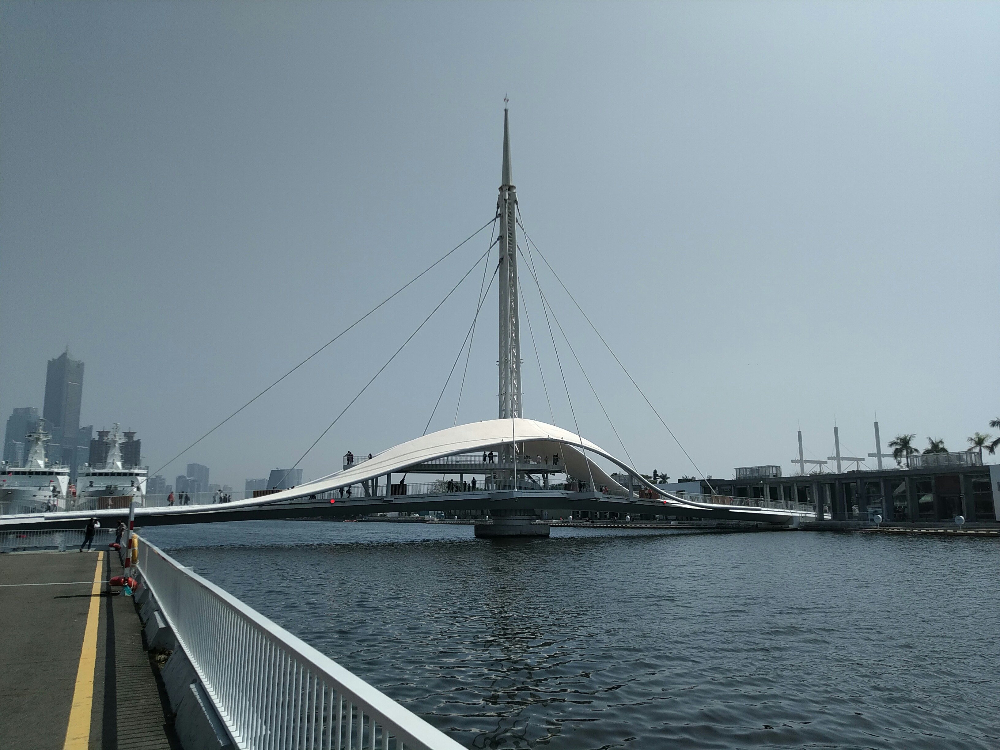 Curved bridge spans a tranquil river with towering skyscrapers in the hazy background.