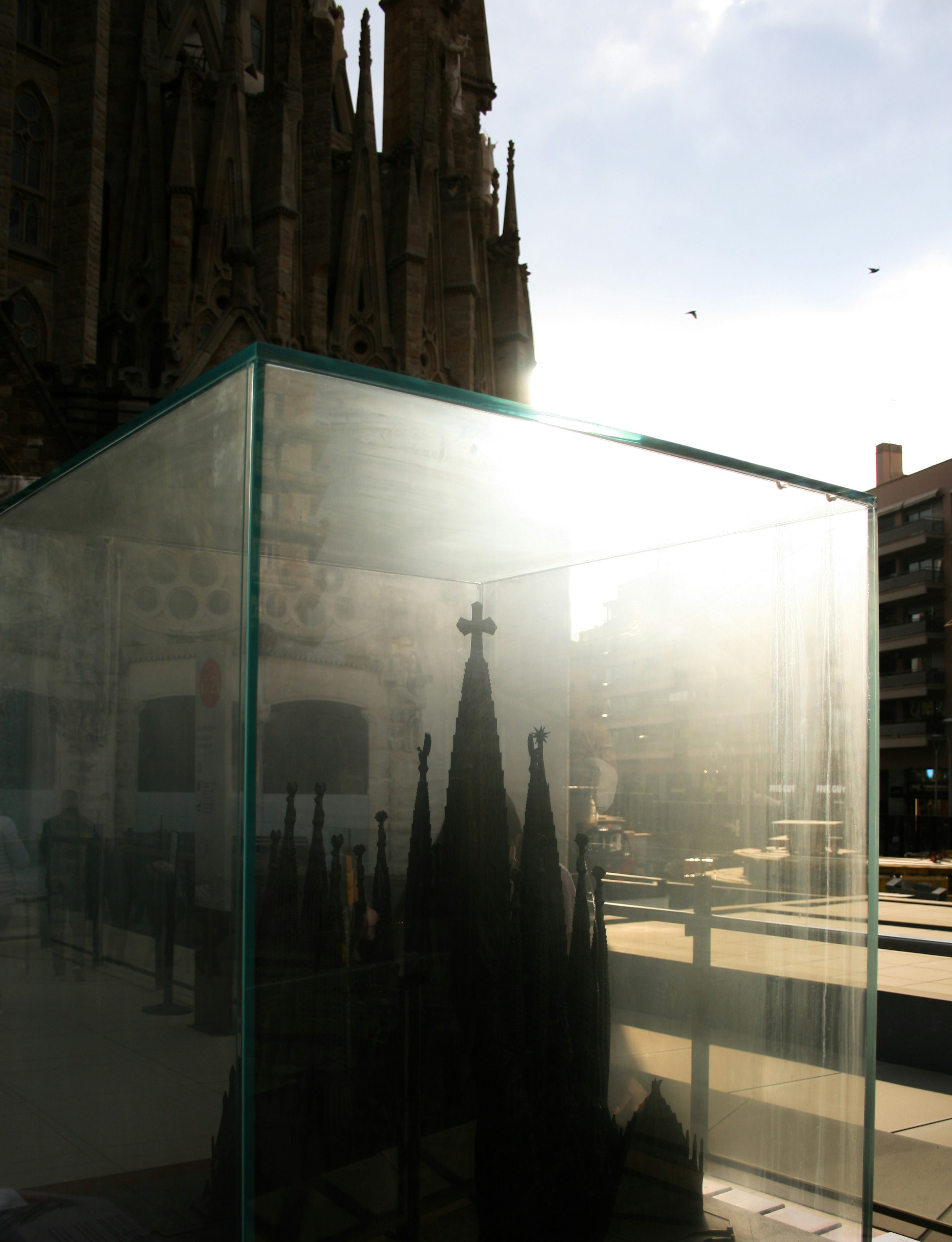 A glass enclosure reveals intricate spires from a historic structure, contrasted against modern architecture and a bright sky.