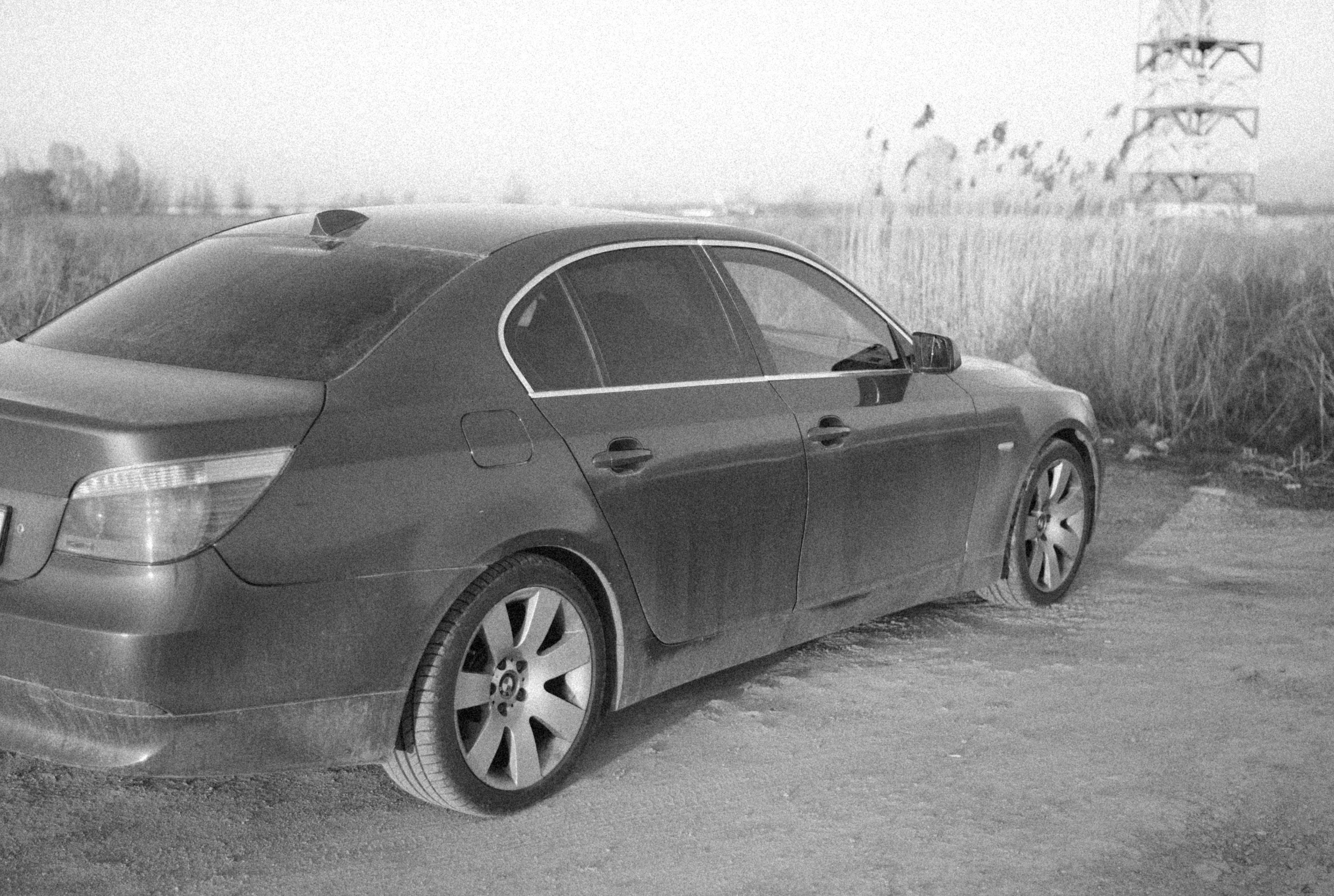 A dusty BMW parked on a gravel path, surrounded by tall grass and power lines in the background.