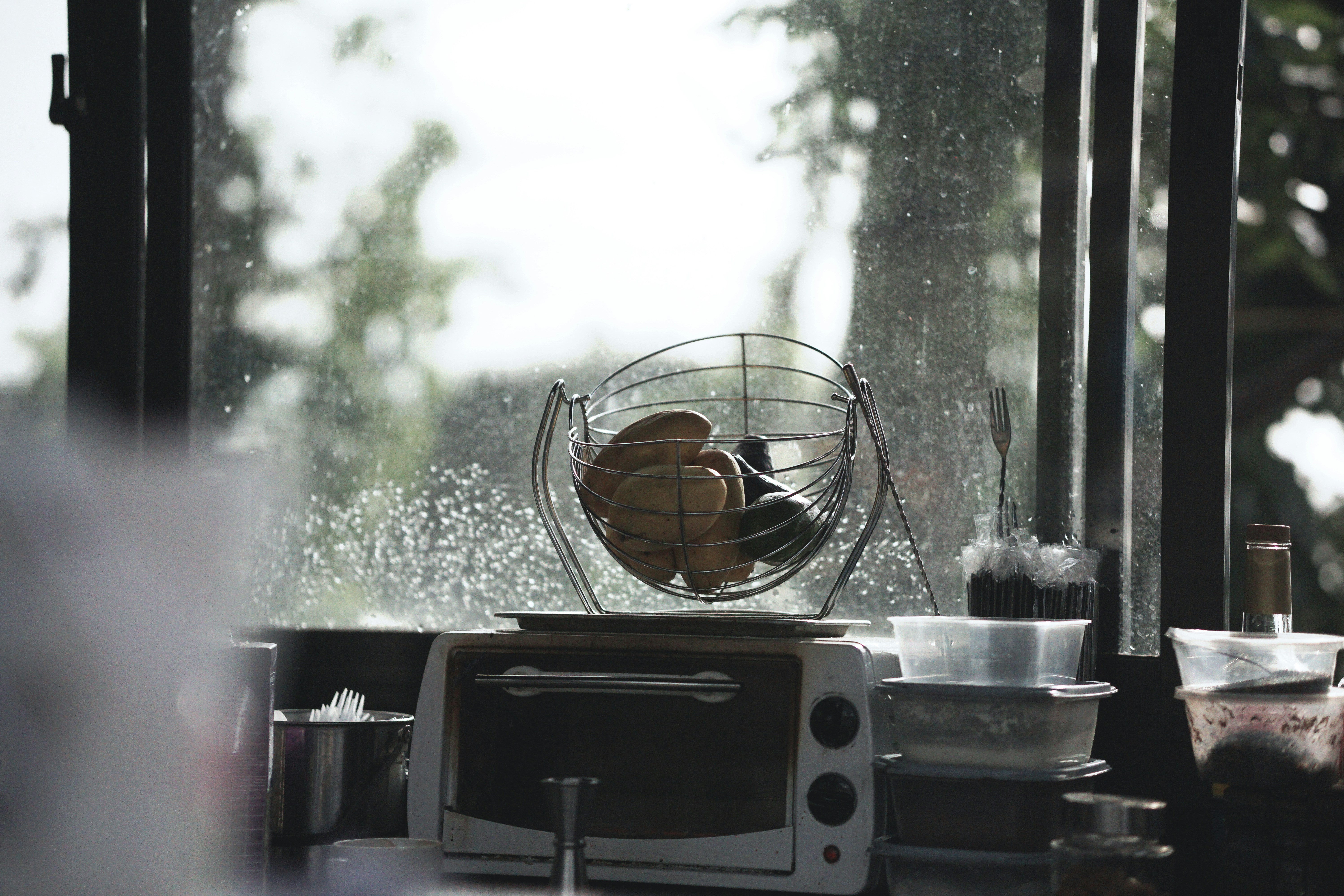 A wire fruit basket filled with bananas and apples sits on a kitchen counter, framed by a rain-speckled window. Soft natural light filters through the glass, enhancing the tranquil atmosphere.