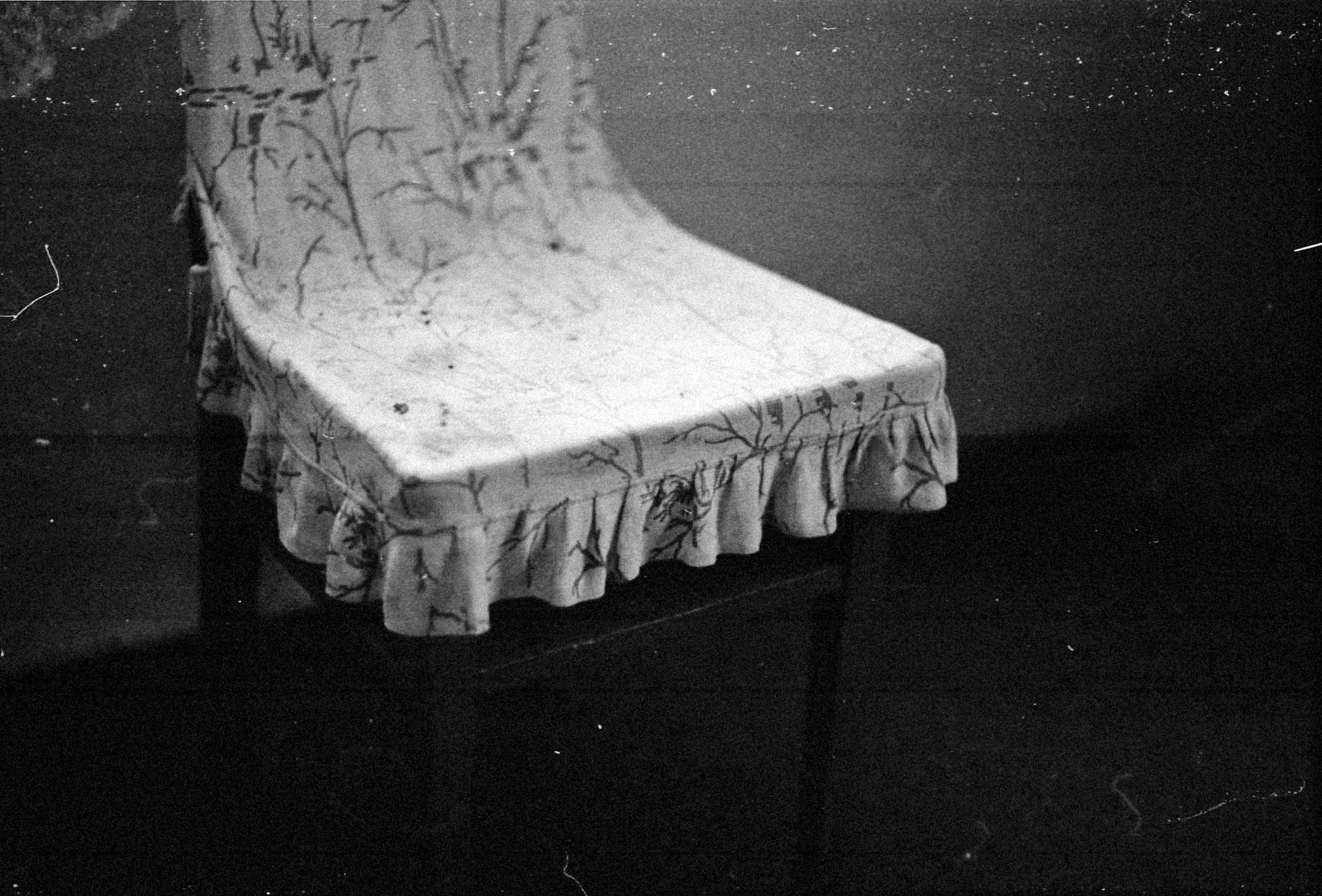 A rickety chair covered with a floral cloth sits in a dim, dusty room, captured in a grainy monochrome that emphasizes texture and age.
