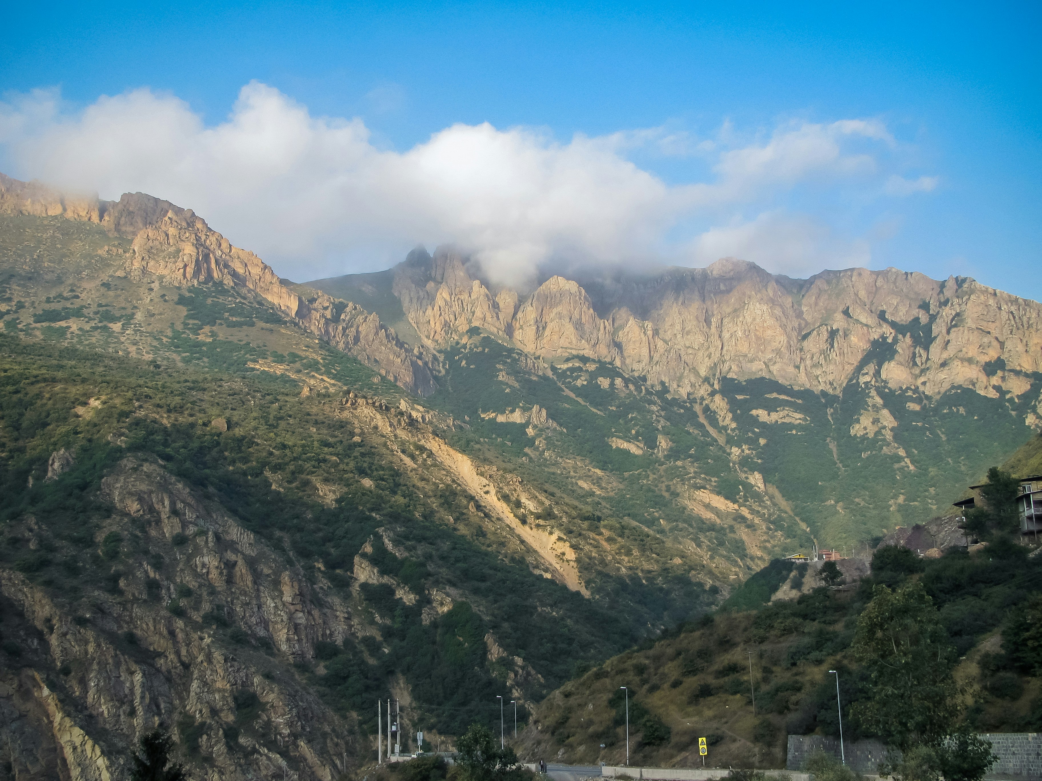 Sunlit limestone ridge towers over a rugged hillside road lined with utility poles and sparse vegetation.
