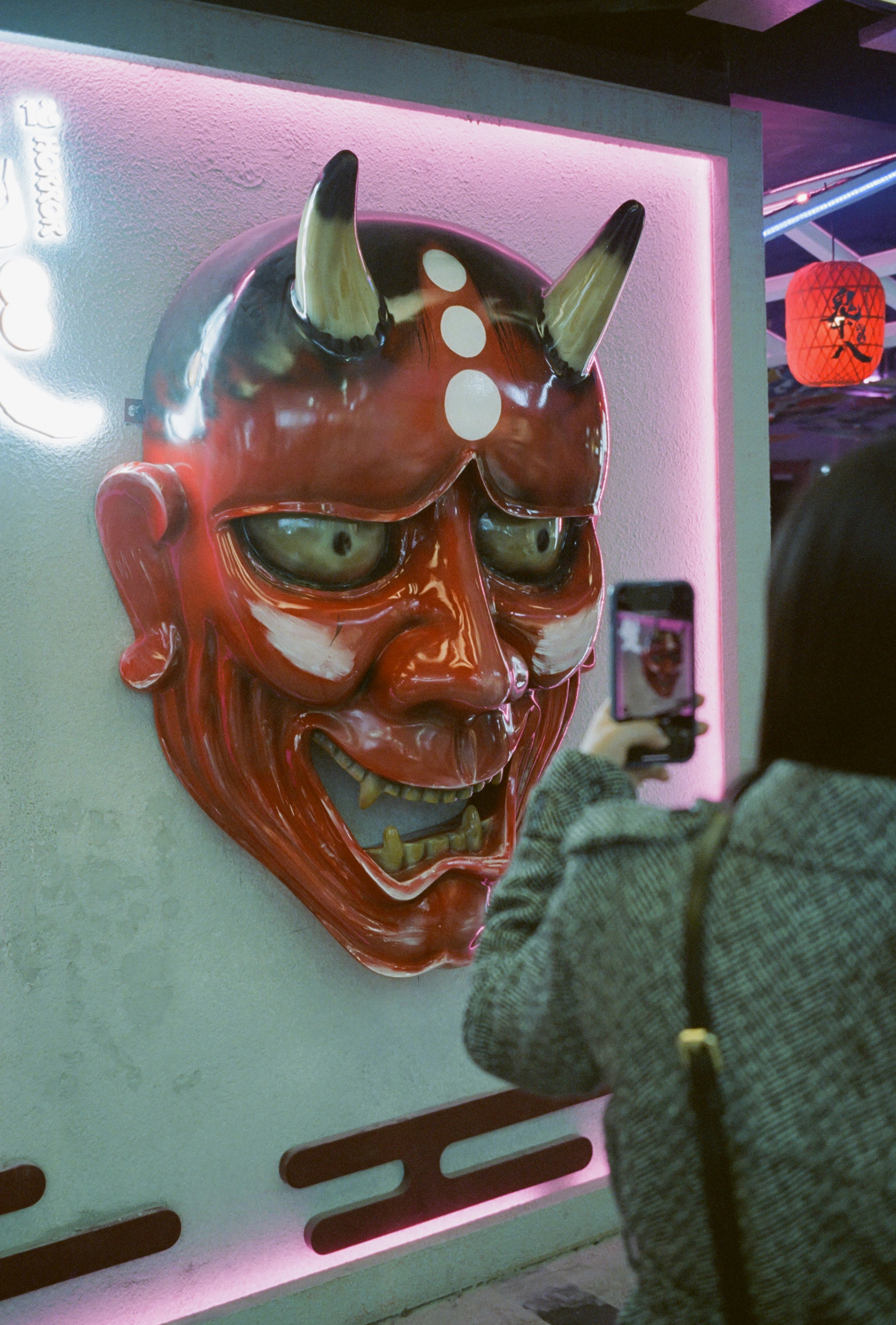 Vibrant red demon mask mounted on a wall, illuminated by soft pink lights, with a person capturing it on their smartphone.