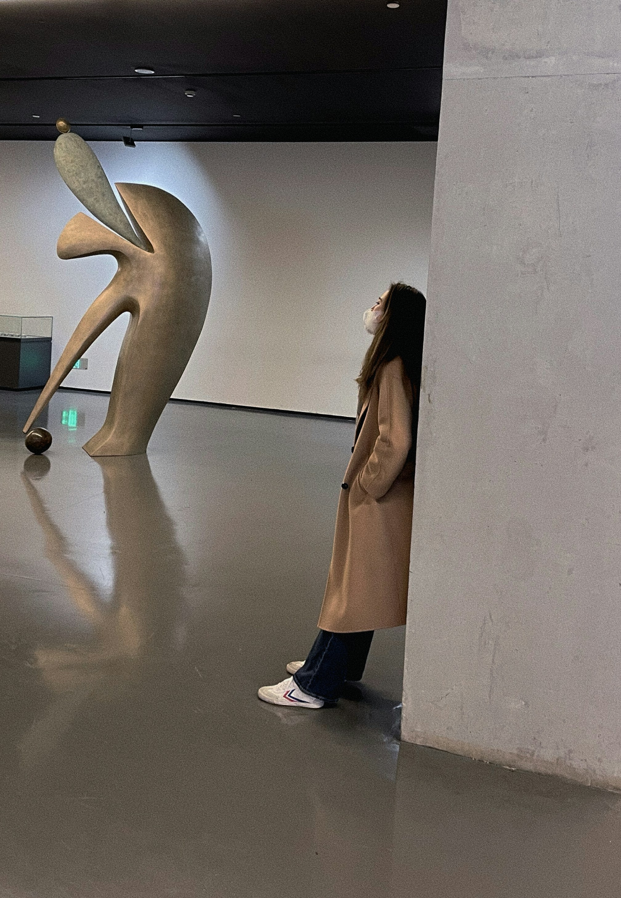 A person leans against a wall, gazing at a modern sculpture in an art gallery. The sleek, reflective floor enhances the minimalist aesthetic.