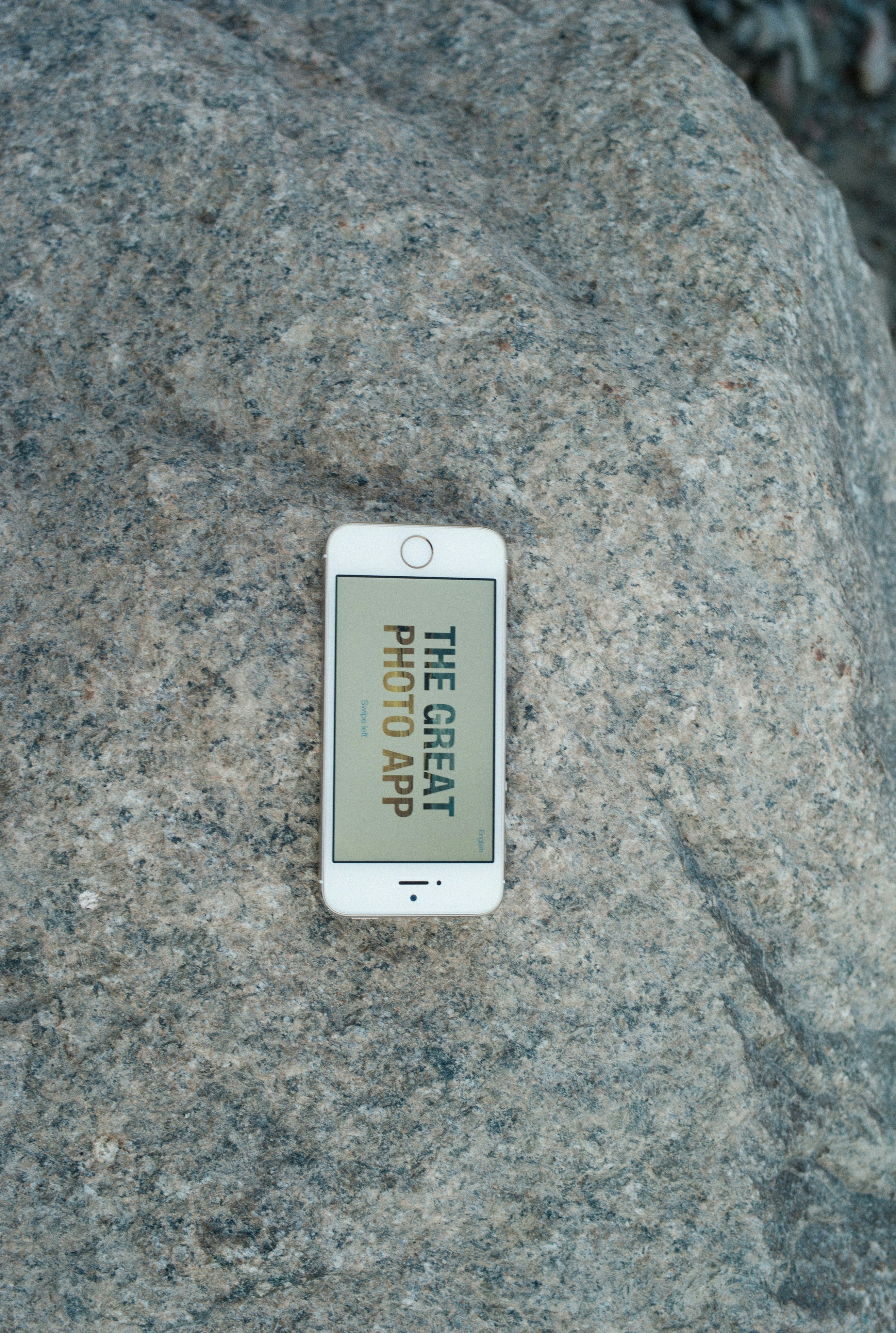 White iPhone rests on textured granite, screen displaying 'THE GREAT PHOTO APP' logo; a minimal scene highlighting branding on a rugged surface.