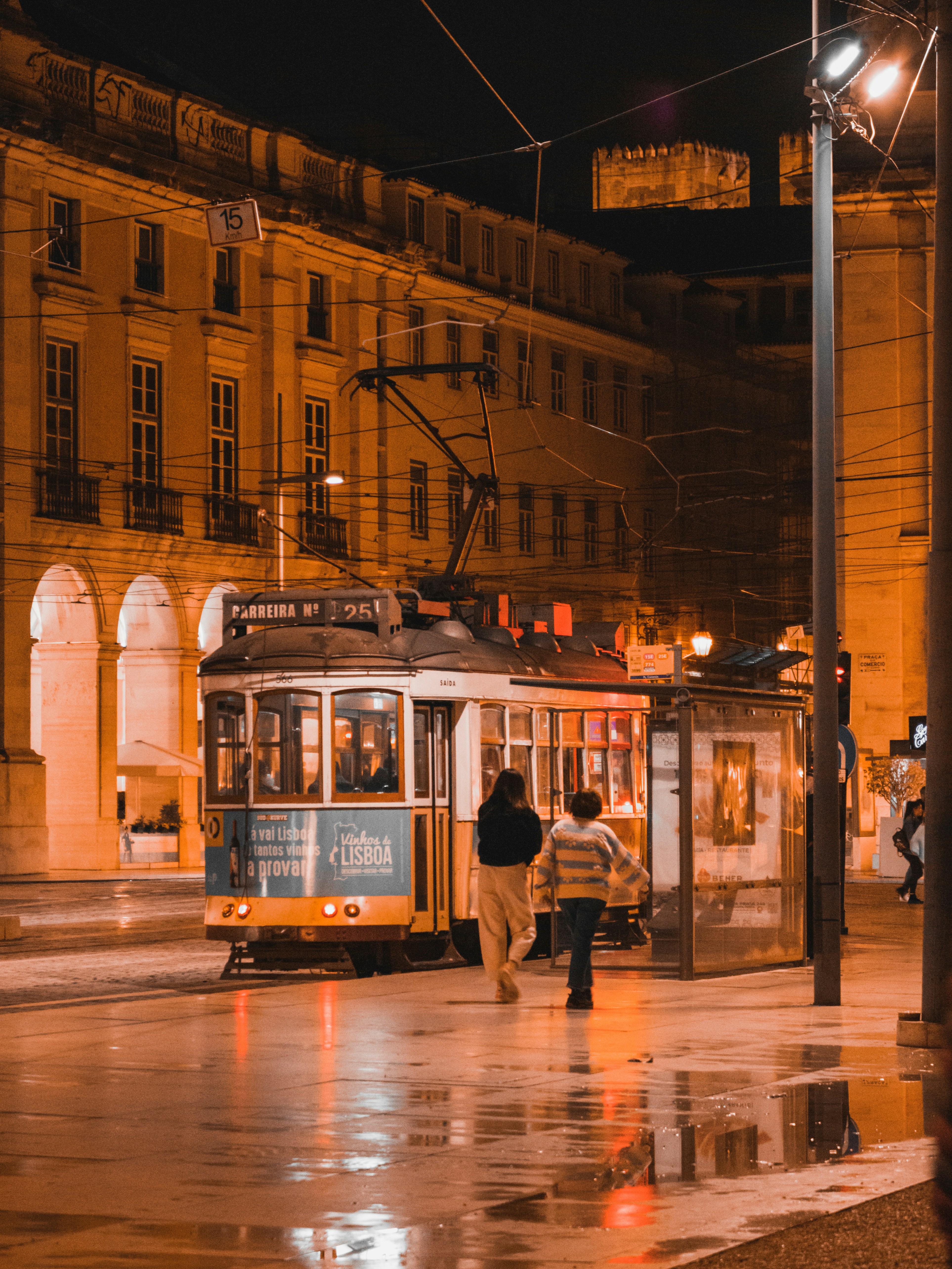 Lisbon tram at night, bathed in orange streetlight tones with reflections on the wet pavement.