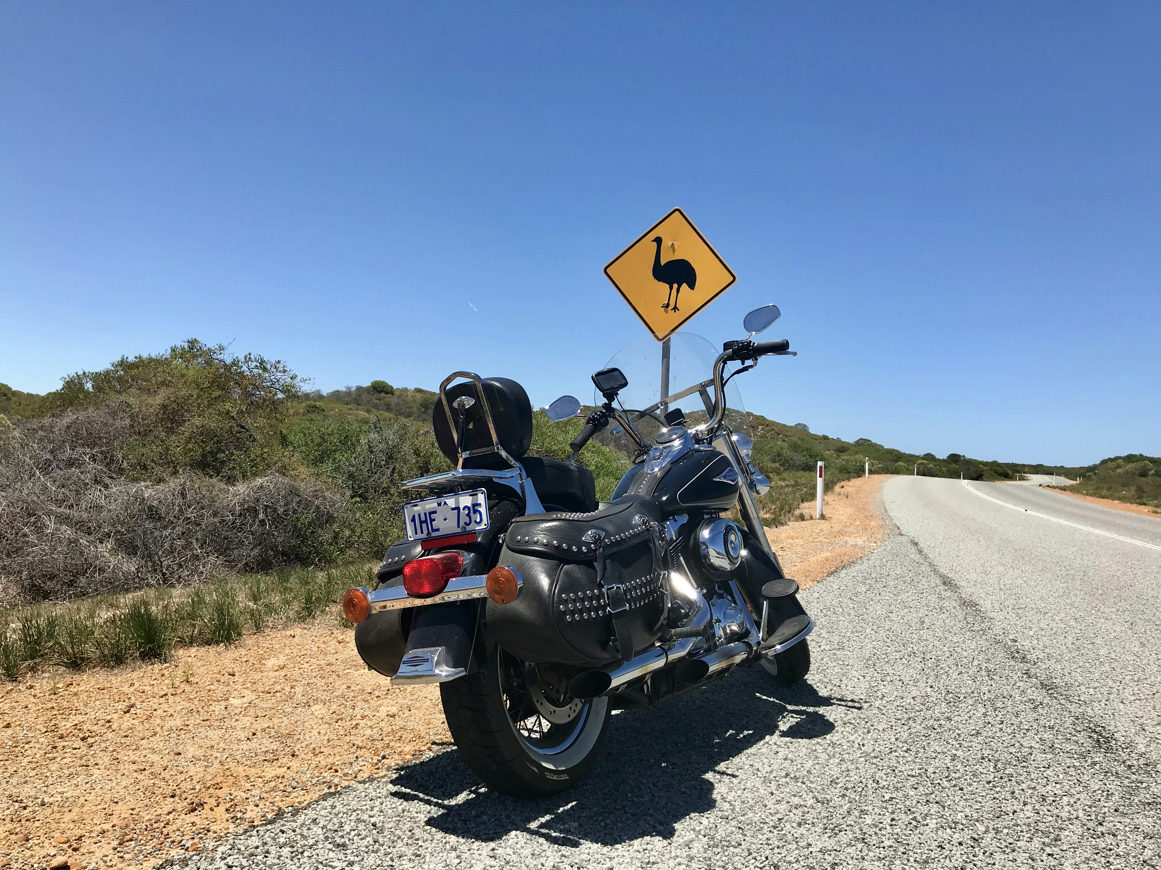 Motorcycle parked beside a road with an ostrich warning sign and a clear blue sky in the background.