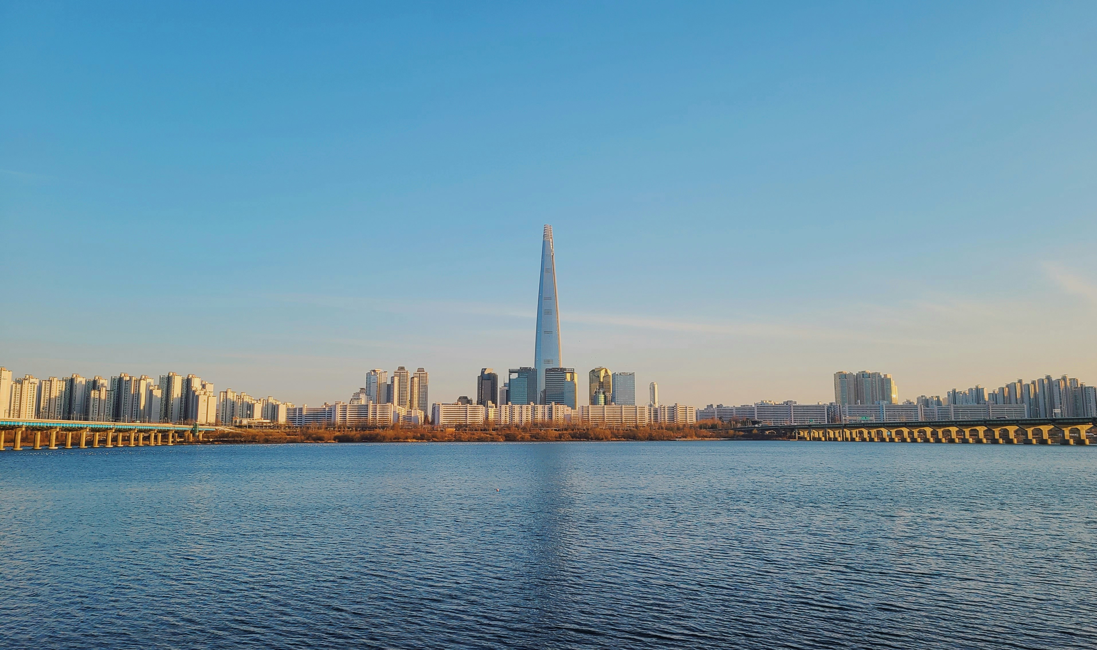 A sleek skyscraper rises above a tranquil river, framed by a sprawling cityscape under a clear blue sky.