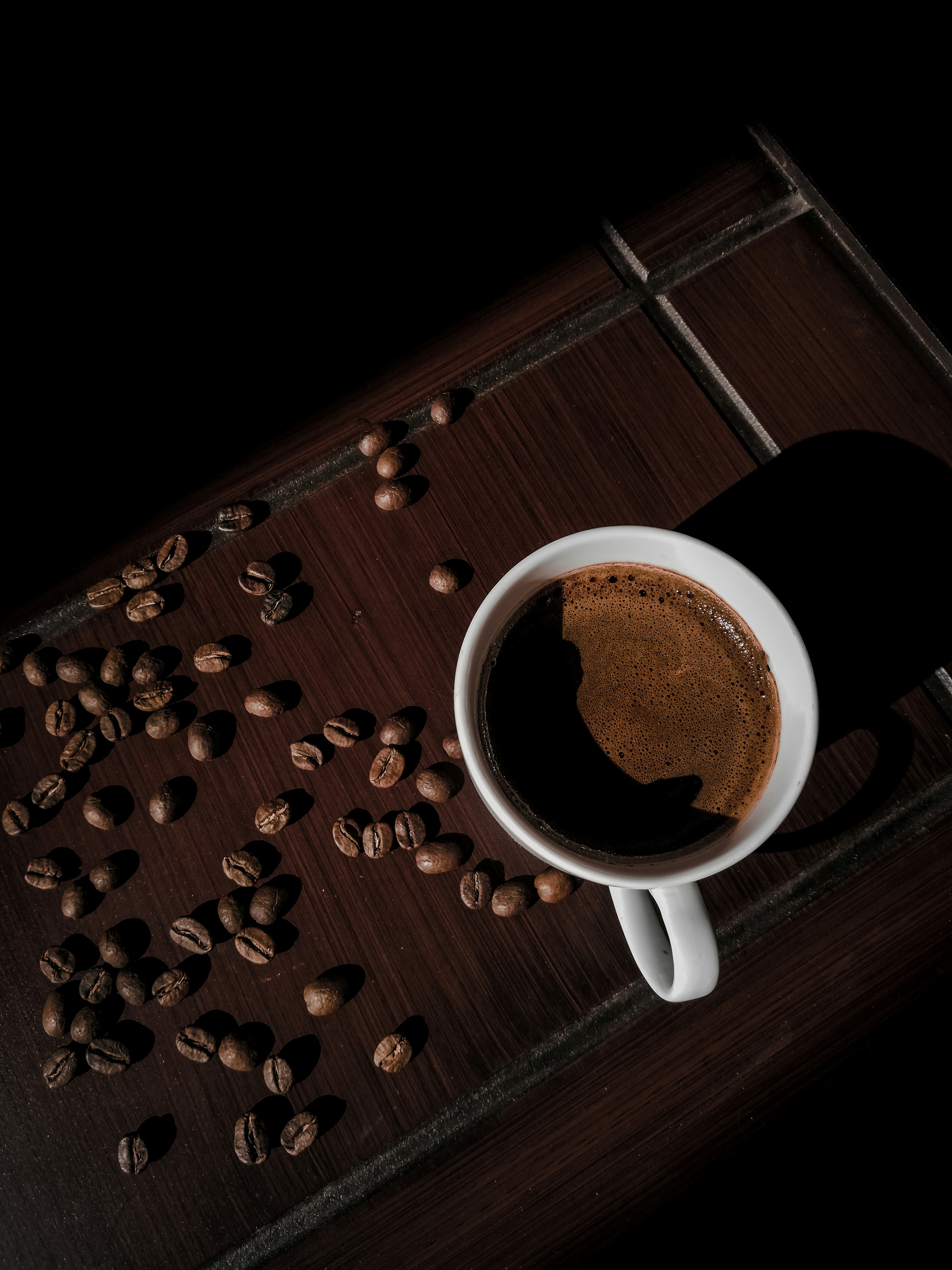 Rich espresso in a white cup beside scattered coffee beans on a dark wooden surface.