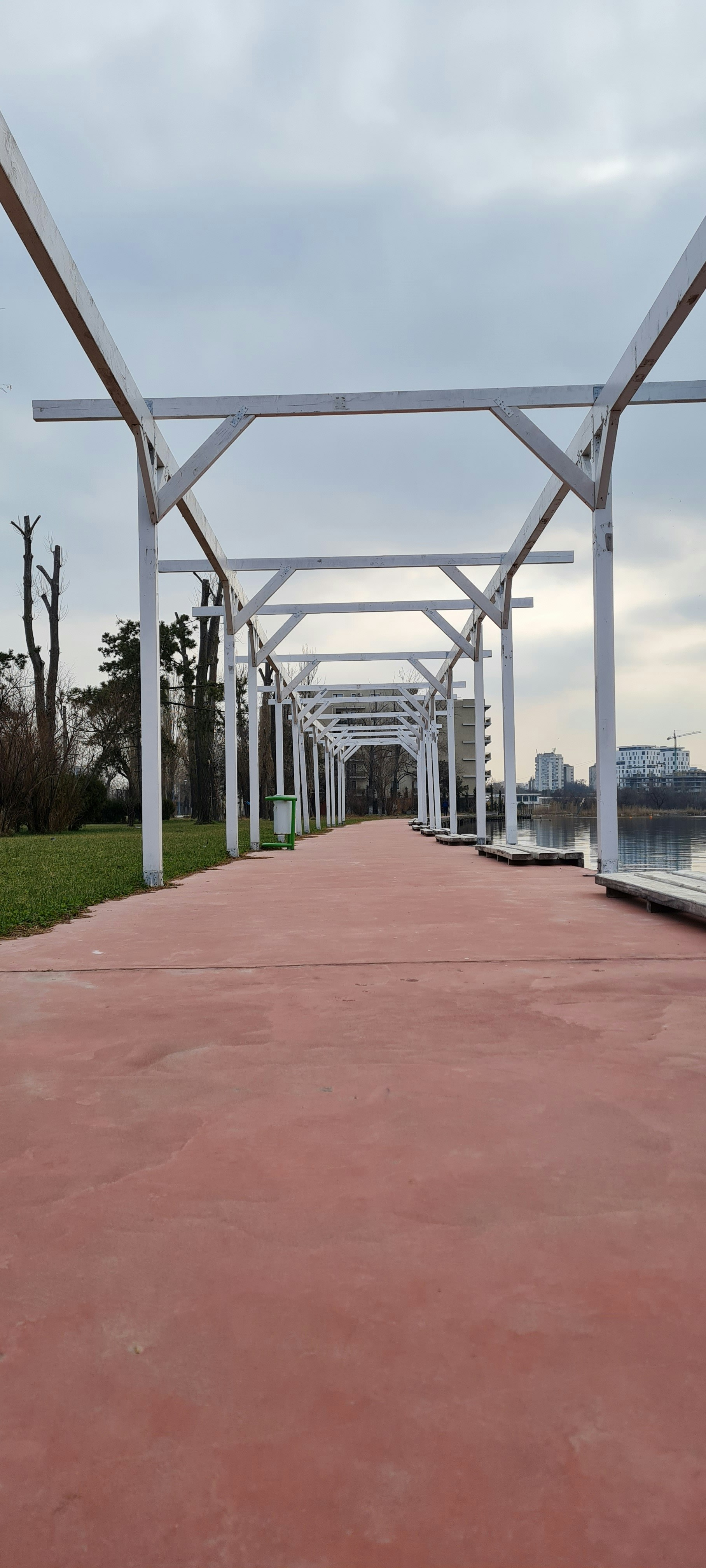 A tranquil walkway framed by white wooden structures beside a calm waterbody, inviting leisurely strolls. The muted colors and soft light create a peaceful atmosphere.