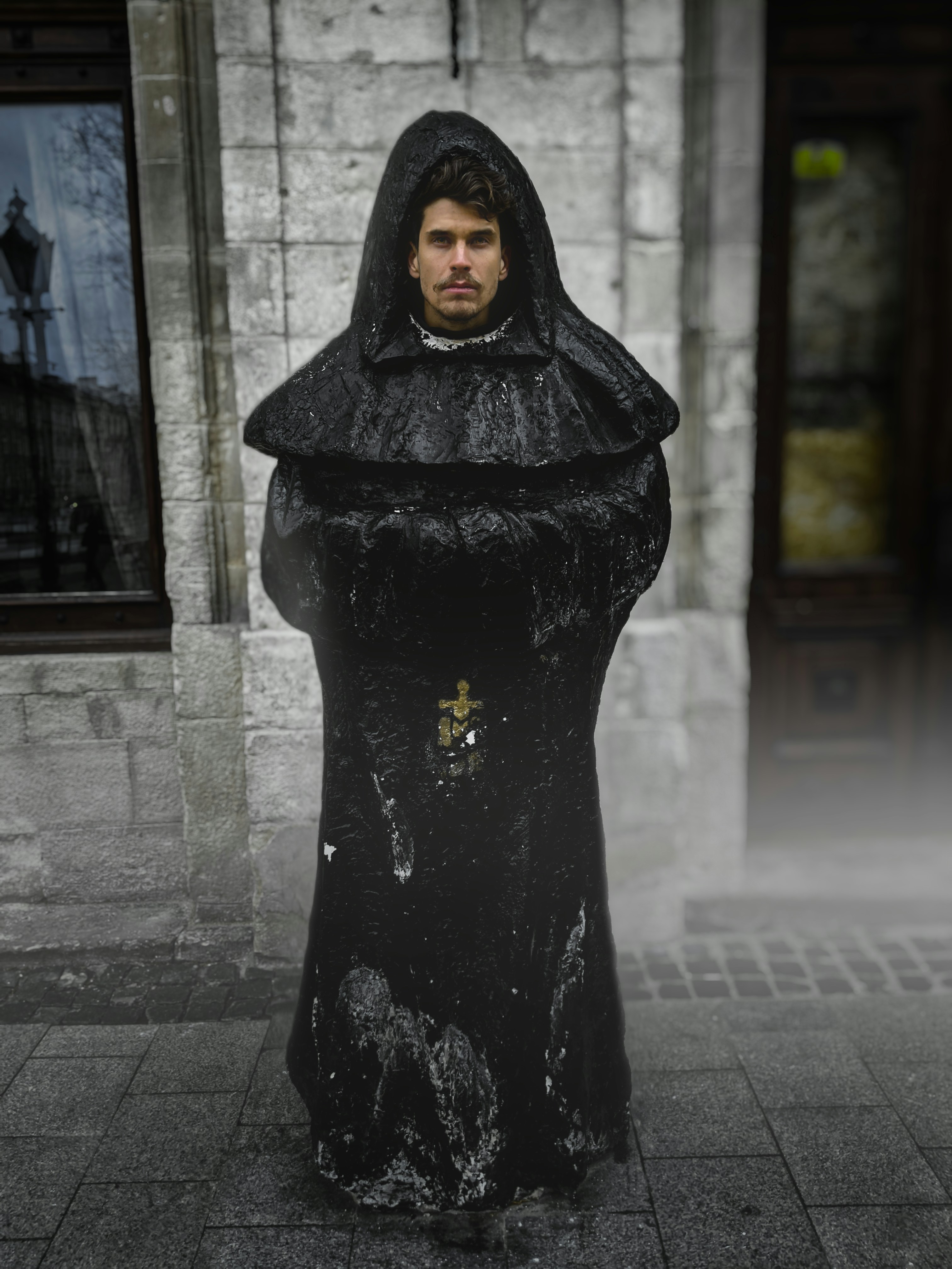 A man dressed as a monk stands still against a textured stone wall, blending into the environment while creating an intriguing visual contrast.