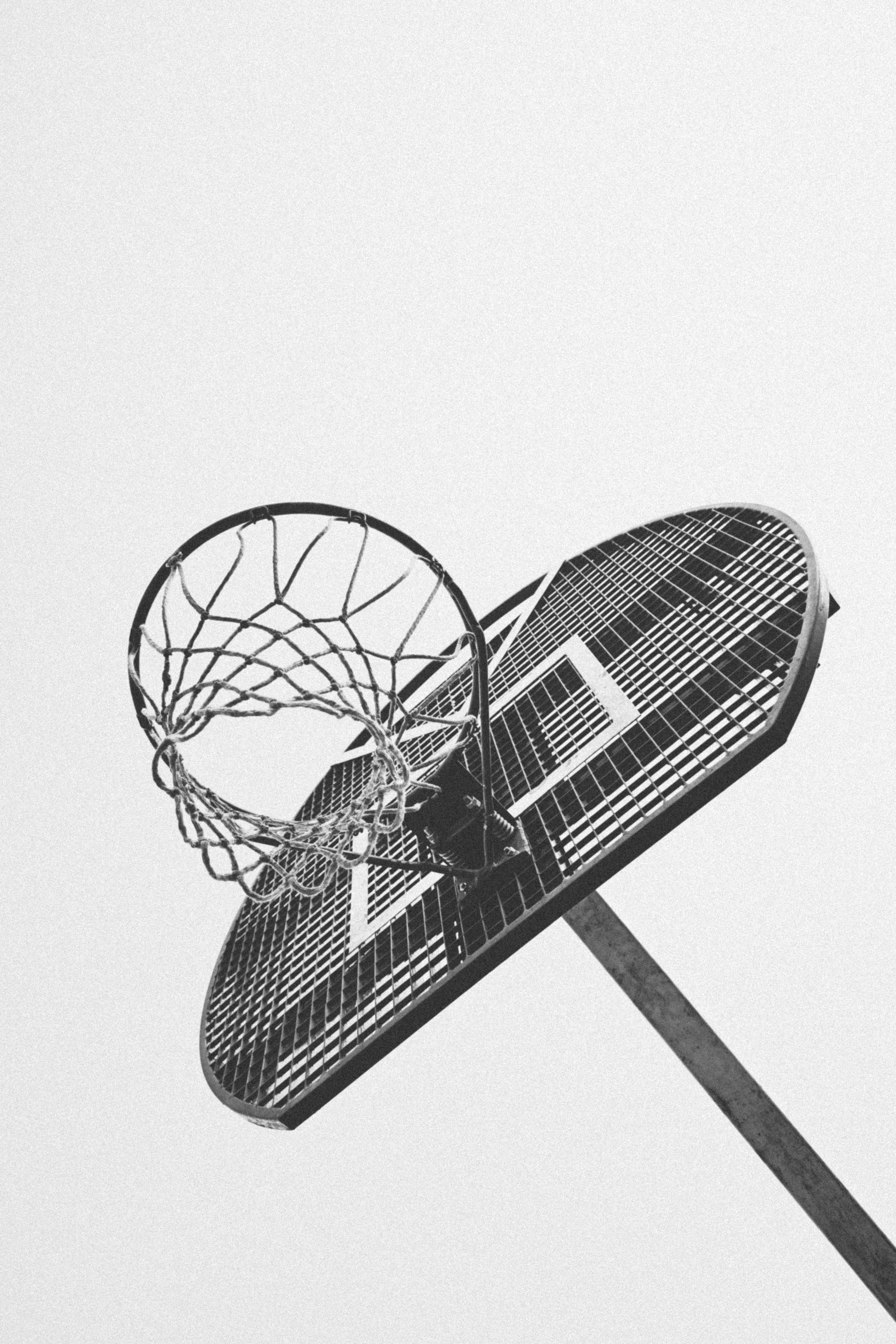 Low-angle view of a basketball hoop against a minimalistic sky, emphasizing the structure and netting. The monochrome tones enhance the graphic quality.