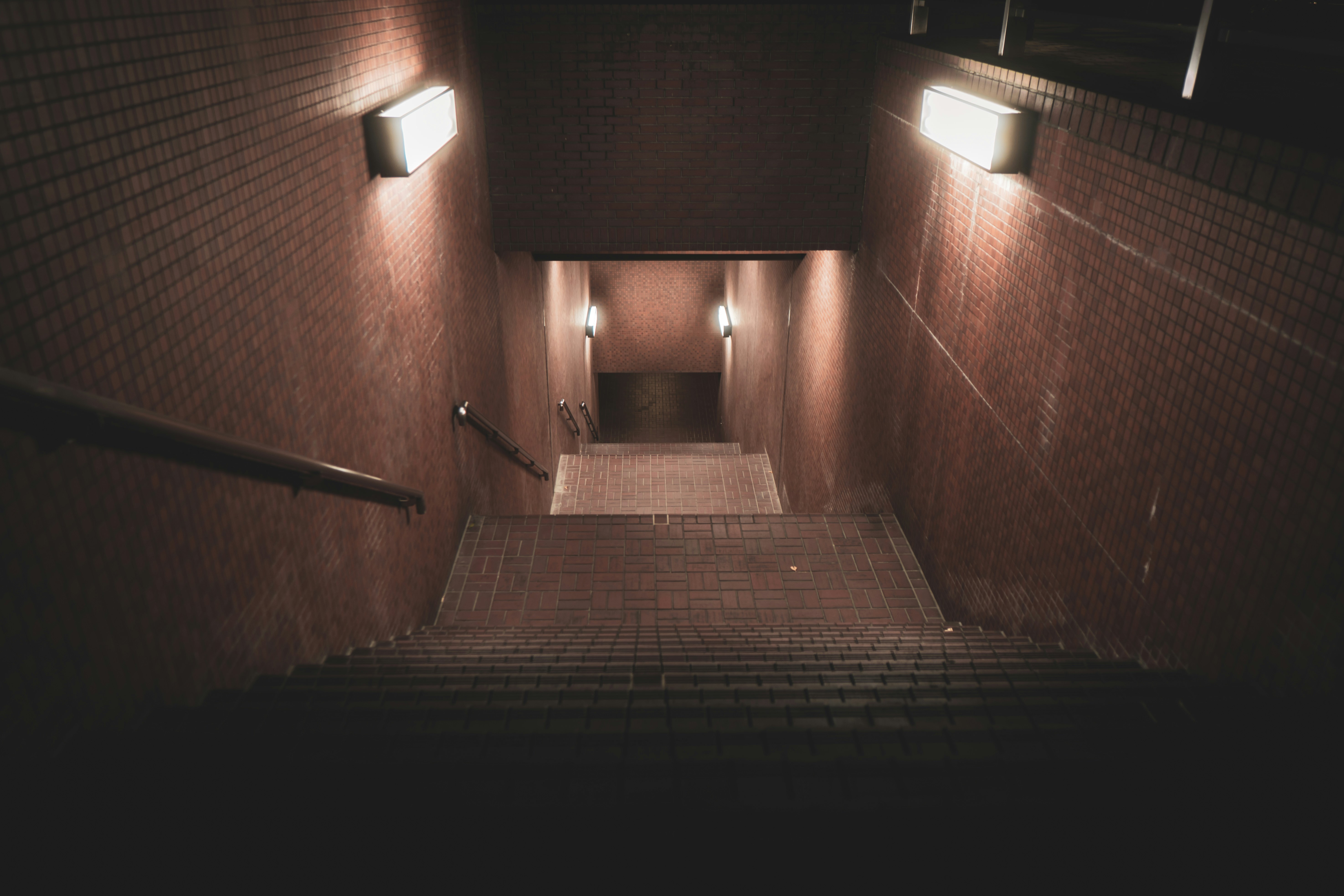 Dimly lit staircase leading down into a dark corridor, illuminated by wall-mounted lights. The textured walls add depth to the scene.