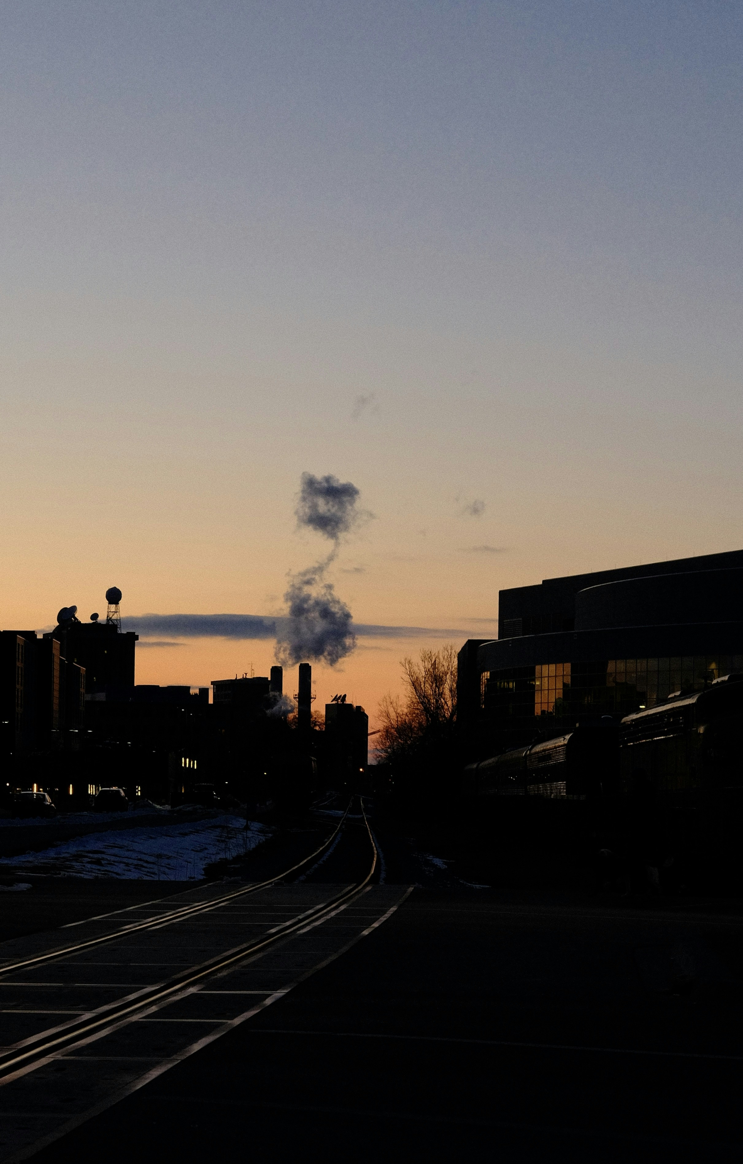 Silhouetted cityscape with steam rising from smokestacks against a twilight sky, highlighting urban life transitioning into night.