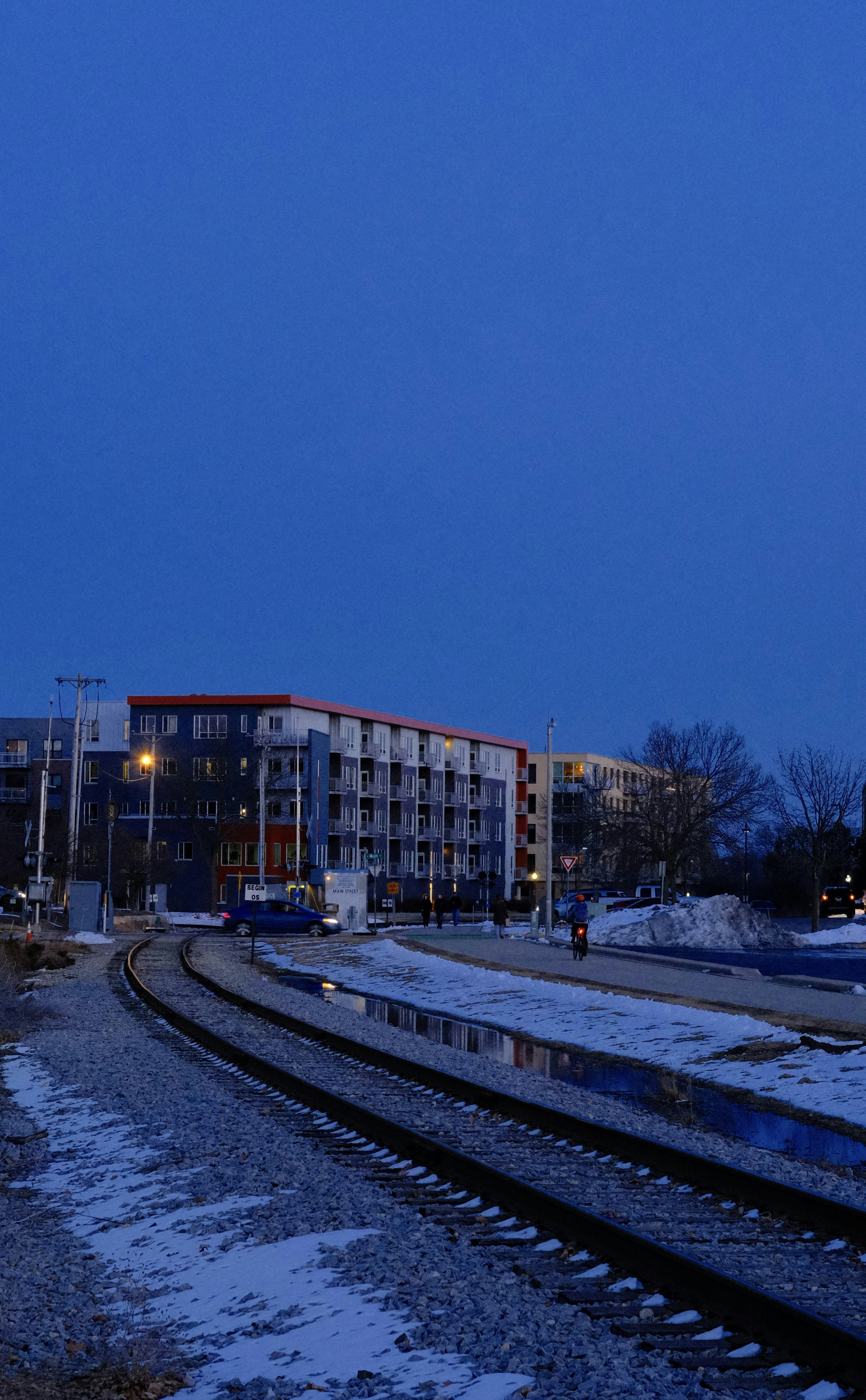 Urban landscape featuring a snow-covered railway with modern buildings under a twilight sky. A solitary figure stands near the tracks.