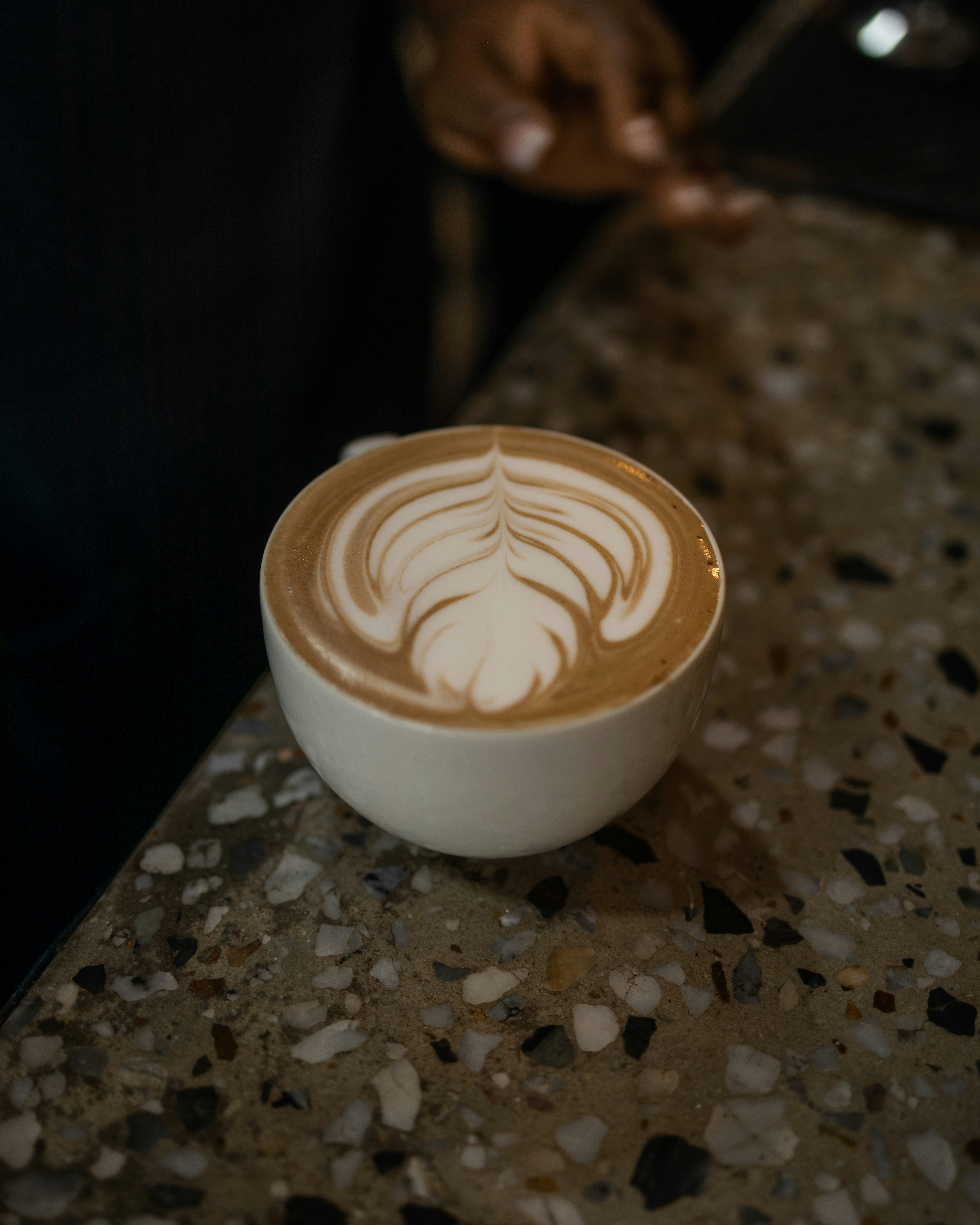 Latte art displayed in a white ceramic cup on a textured stone countertop, showcasing intricate patterns. A hand is subtly visible, adding a human touch to the scene.