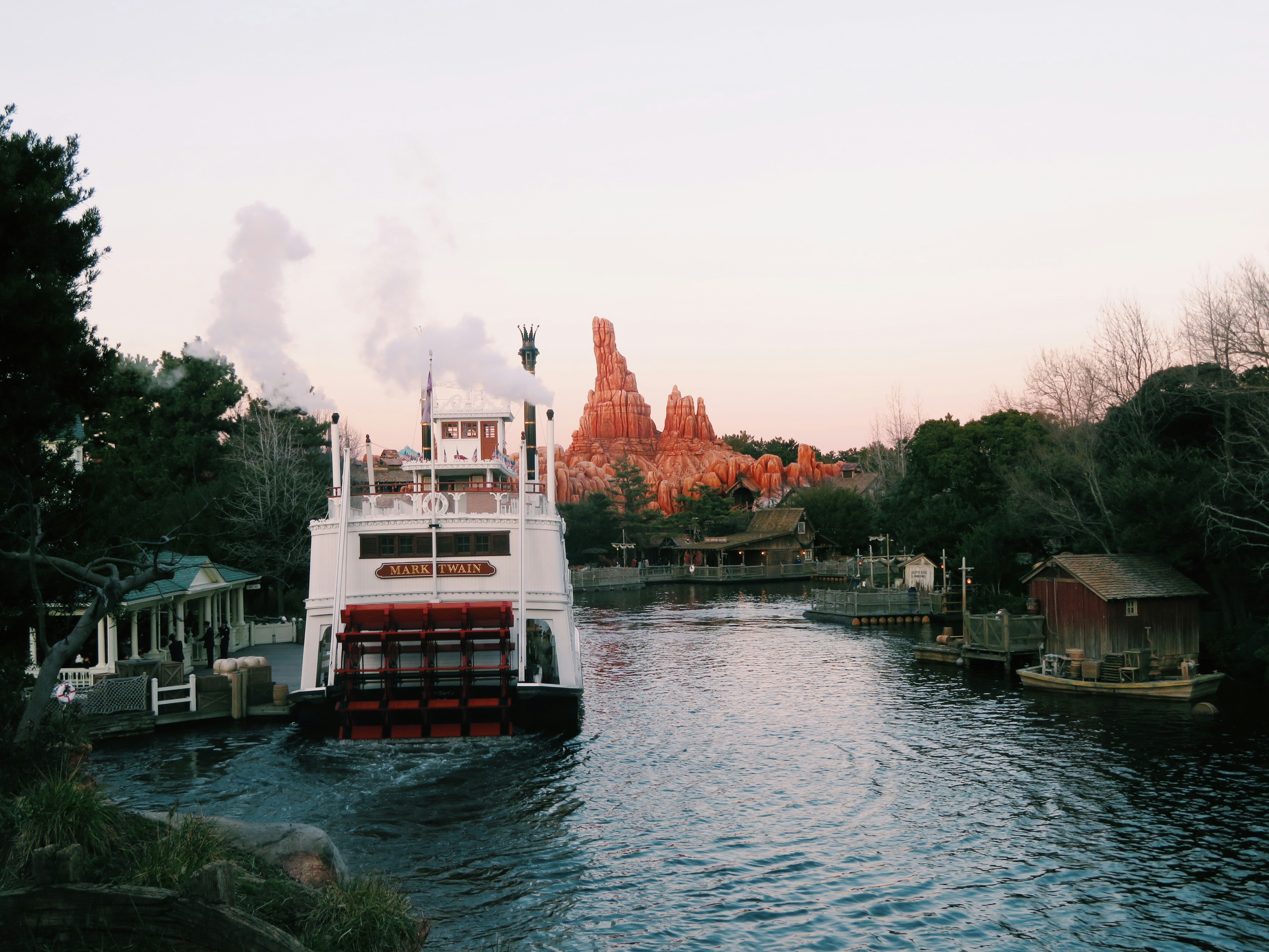 Large riverboat cruising along a tree-lined river with distant rocky formations at sunset.