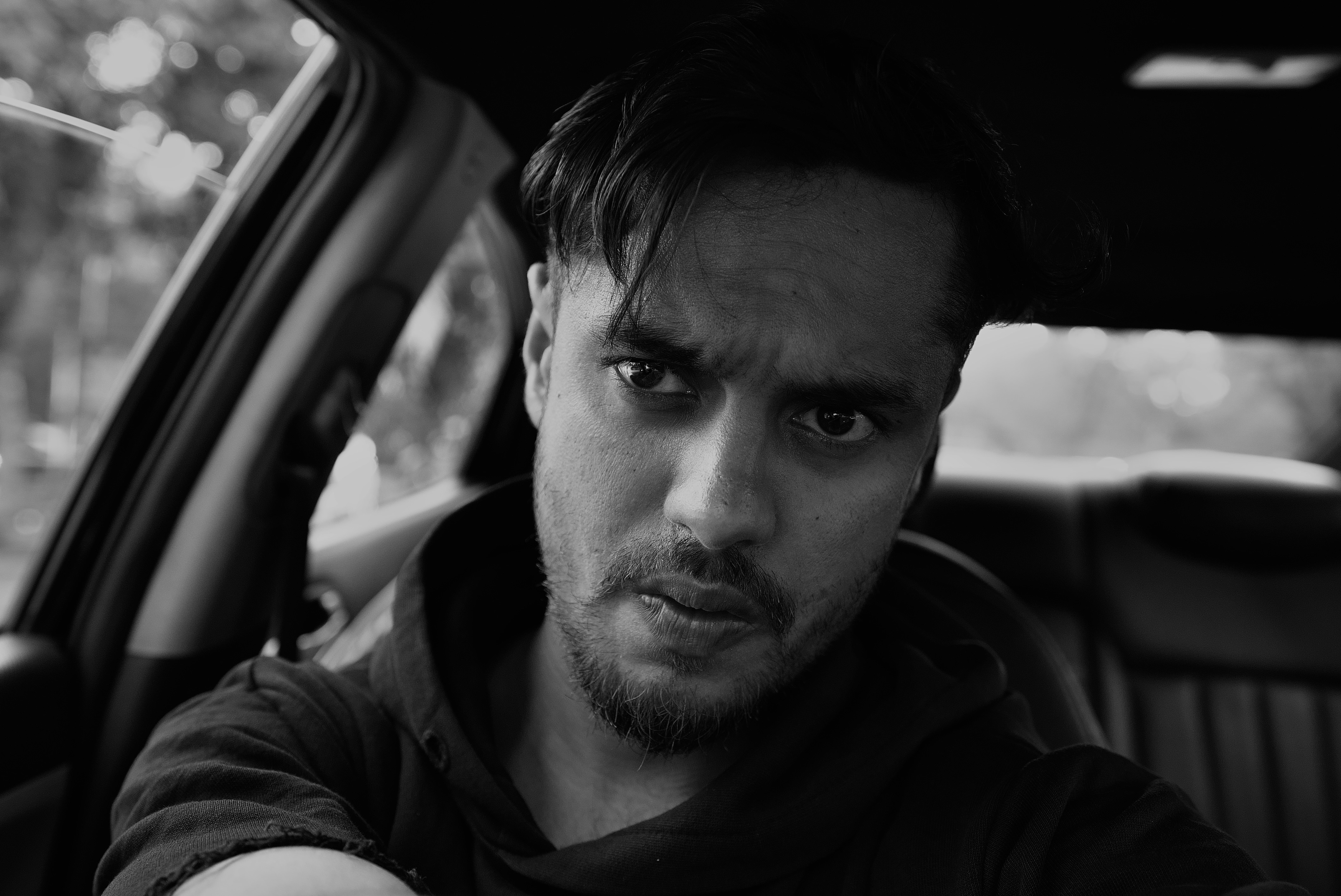 Black and white portrait of a person sitting inside a car, looking directly at the camera.