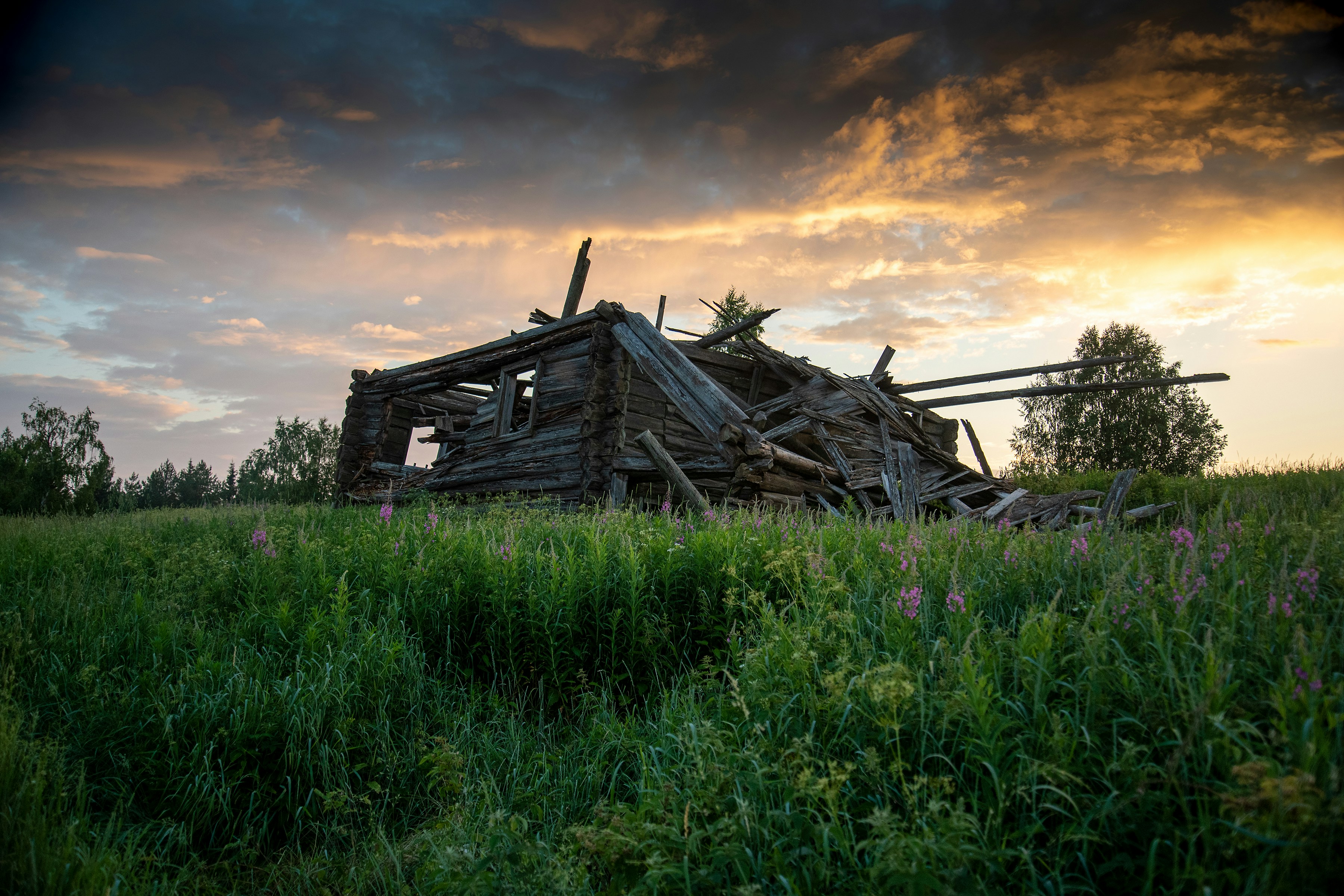 Dilapidated wooden structure in a lush green field under a dramatic sunset sky.