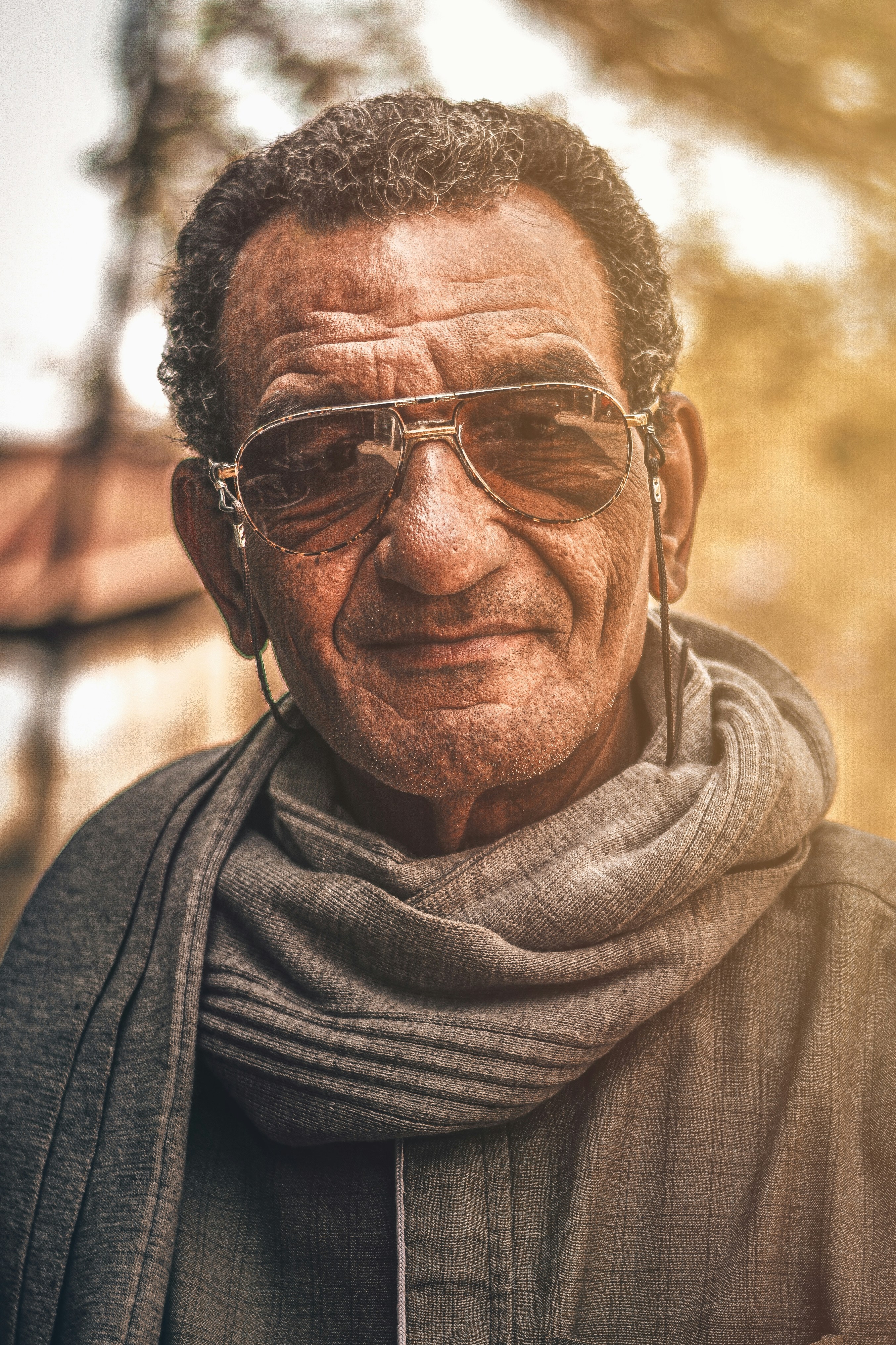 Elderly man wearing sunglasses and a scarf, exuding a sense of wisdom and calmness. The warm background hints at a serene environment.